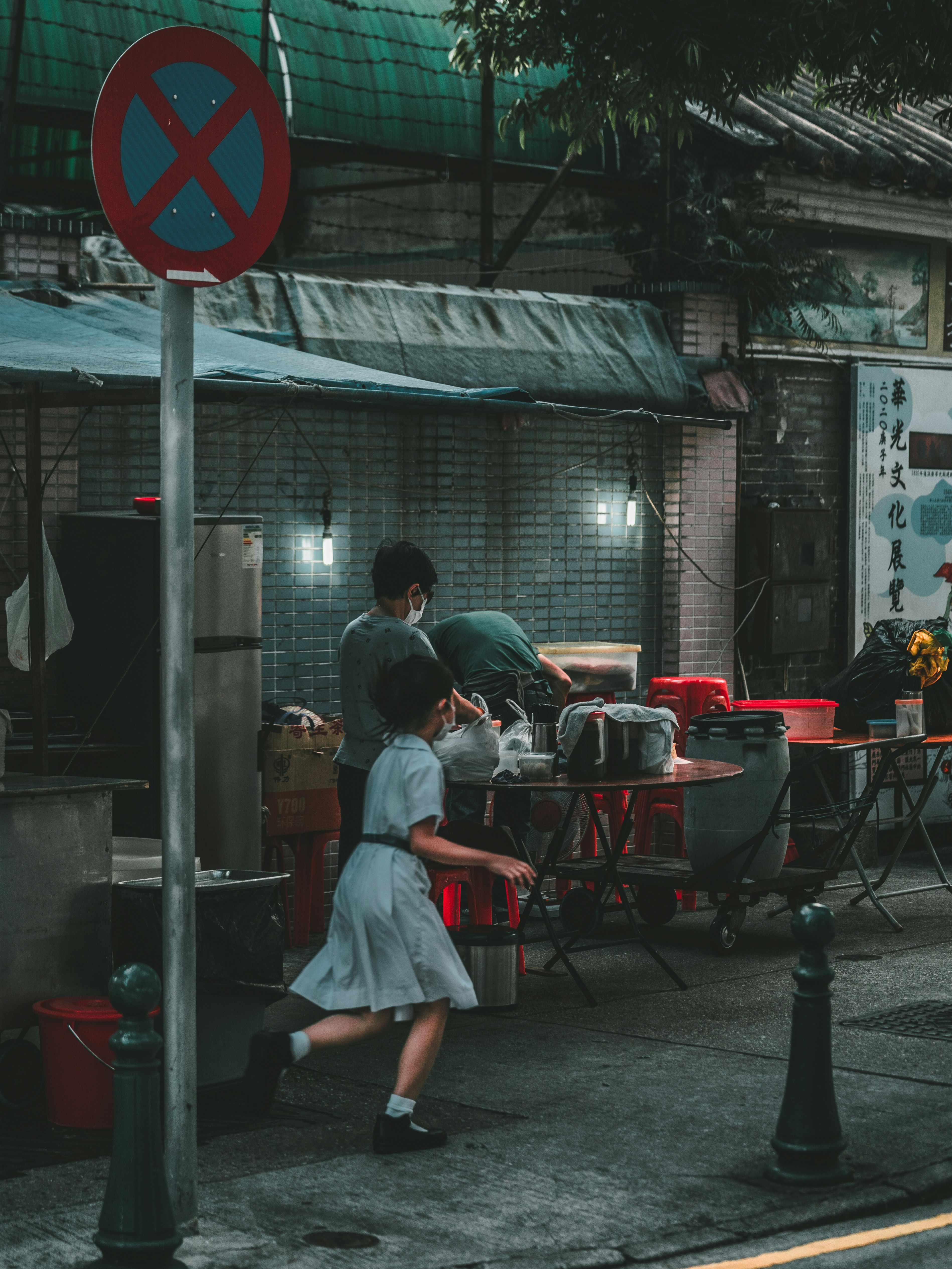A young girl in a school uniform dashes past a bustling street food stall, where vendors prepare meals under soft lighting. The scene captures the essence of urban daily life.