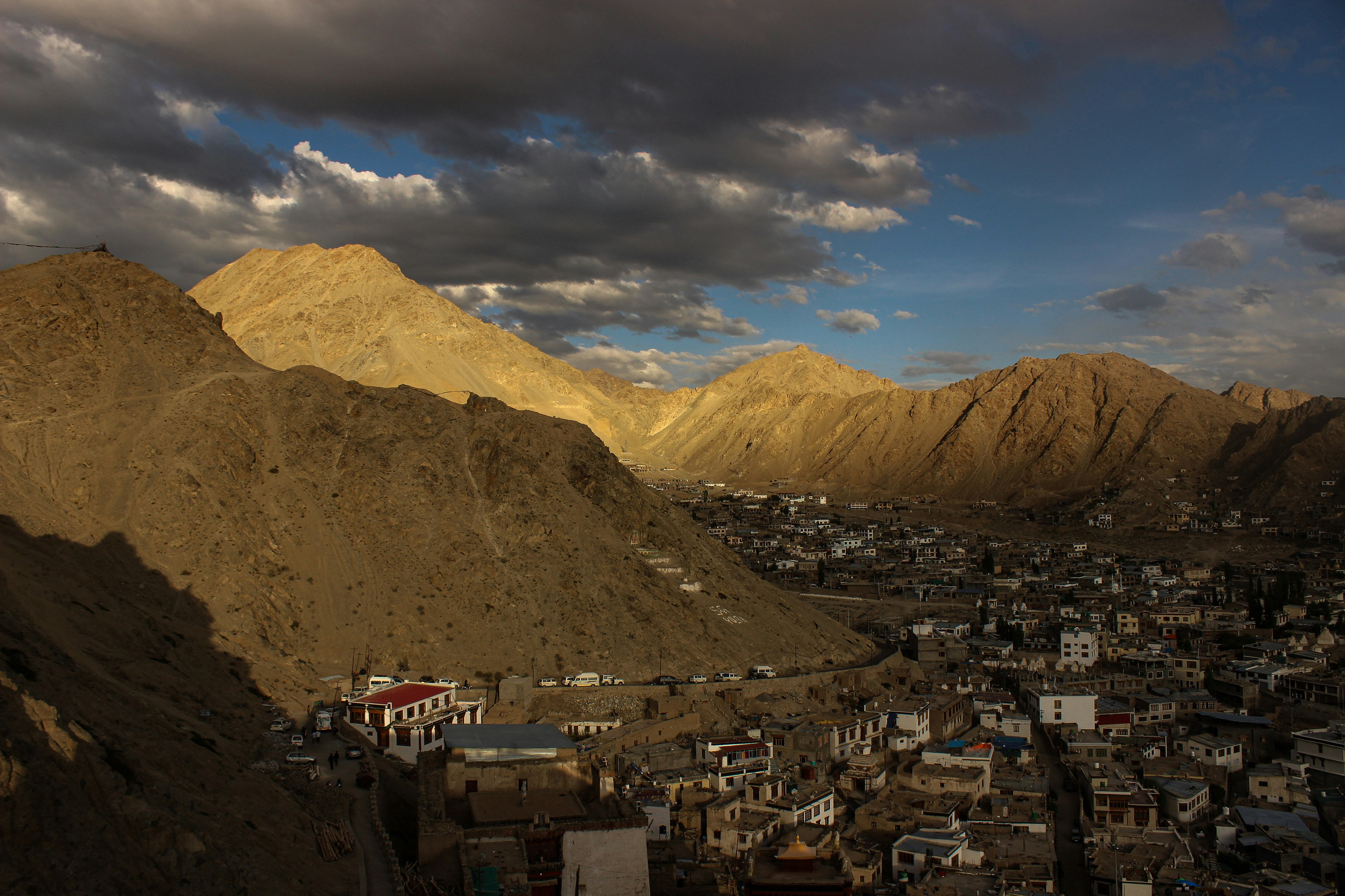 Aerial view of a village nestled in a valley surrounded by rugged mountains under a dramatic sky. The interplay of light and shadow highlights the terrain's textures.
