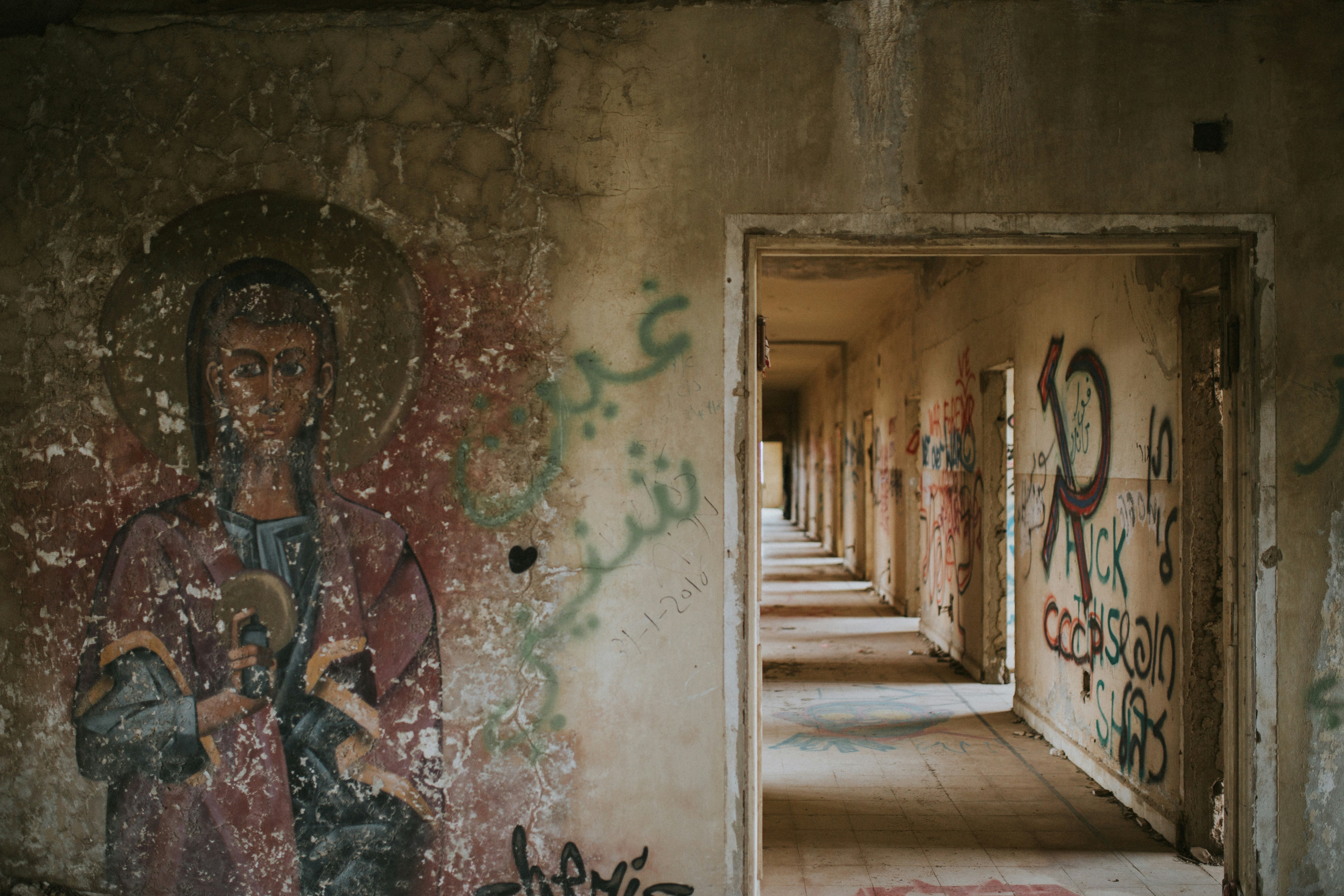 Faded mural of a saint juxtaposed against graffiti in an abandoned corridor. The scene captures the contrast between art and decay.