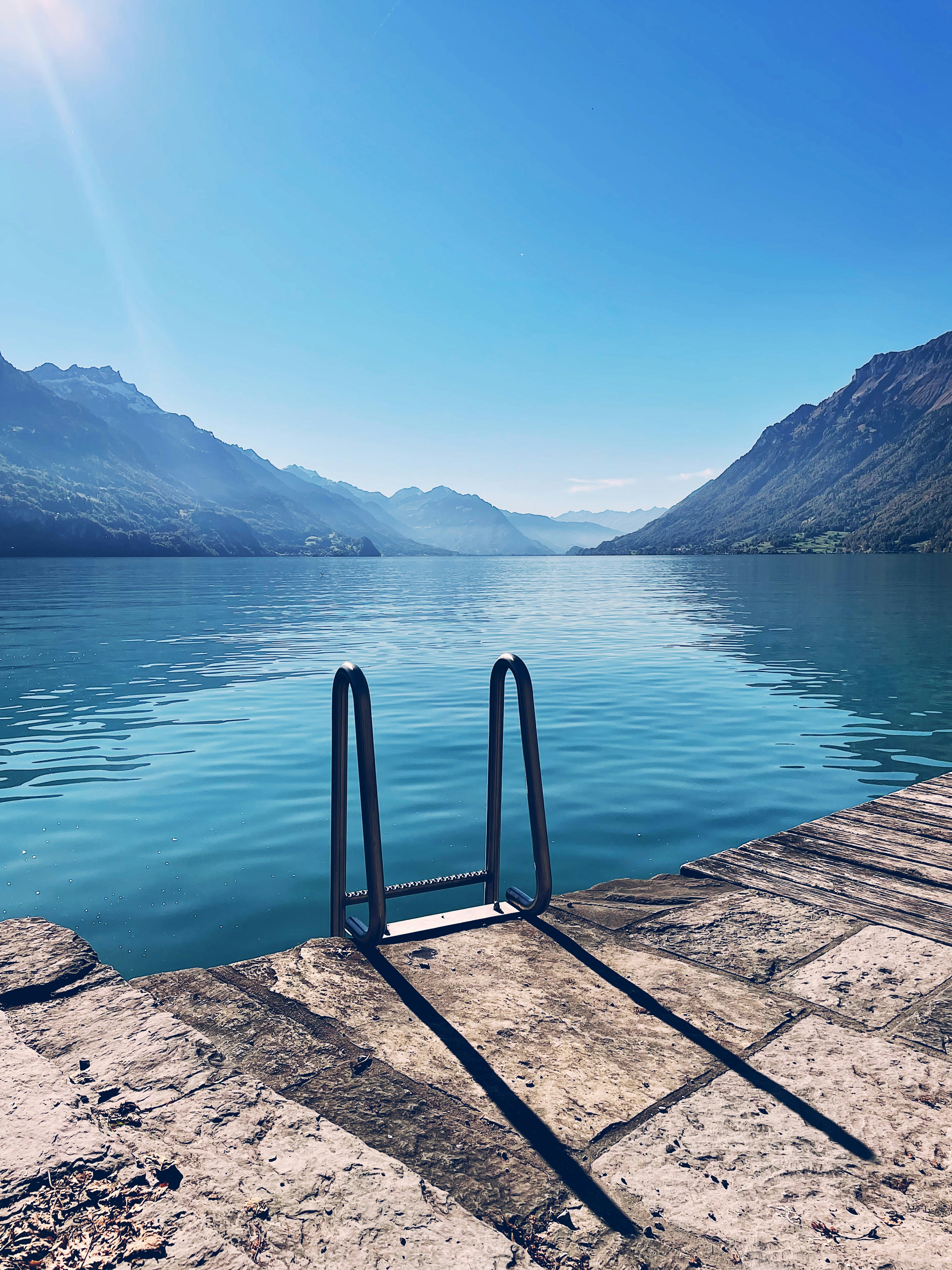 Metal ladder leading into serene blue lake surrounded by mountains under clear skies.