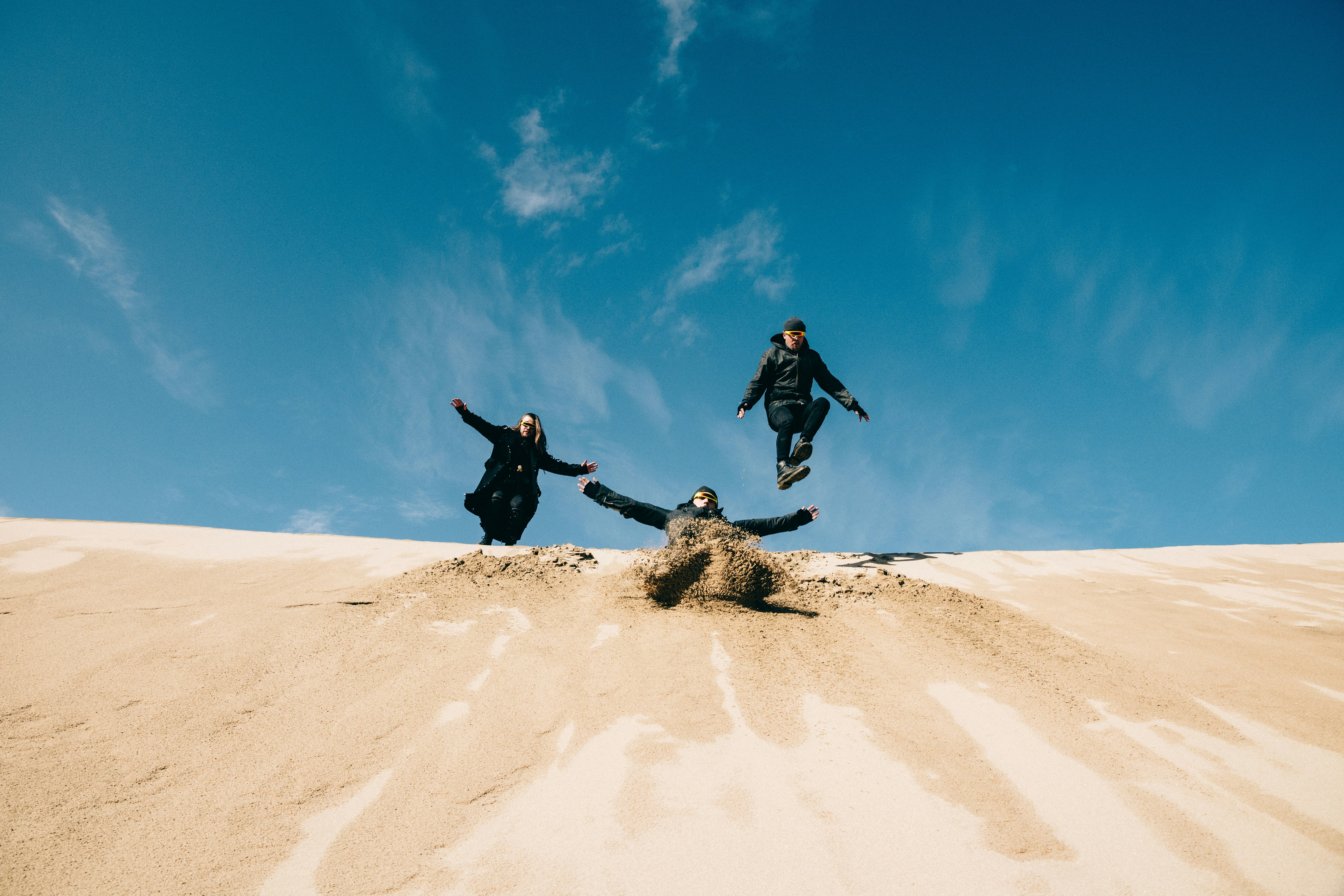 Three friends joyfully playing on a sandy dune, with one mid-air jump and two others in playful poses. The bright blue sky contrasts with the golden sand.