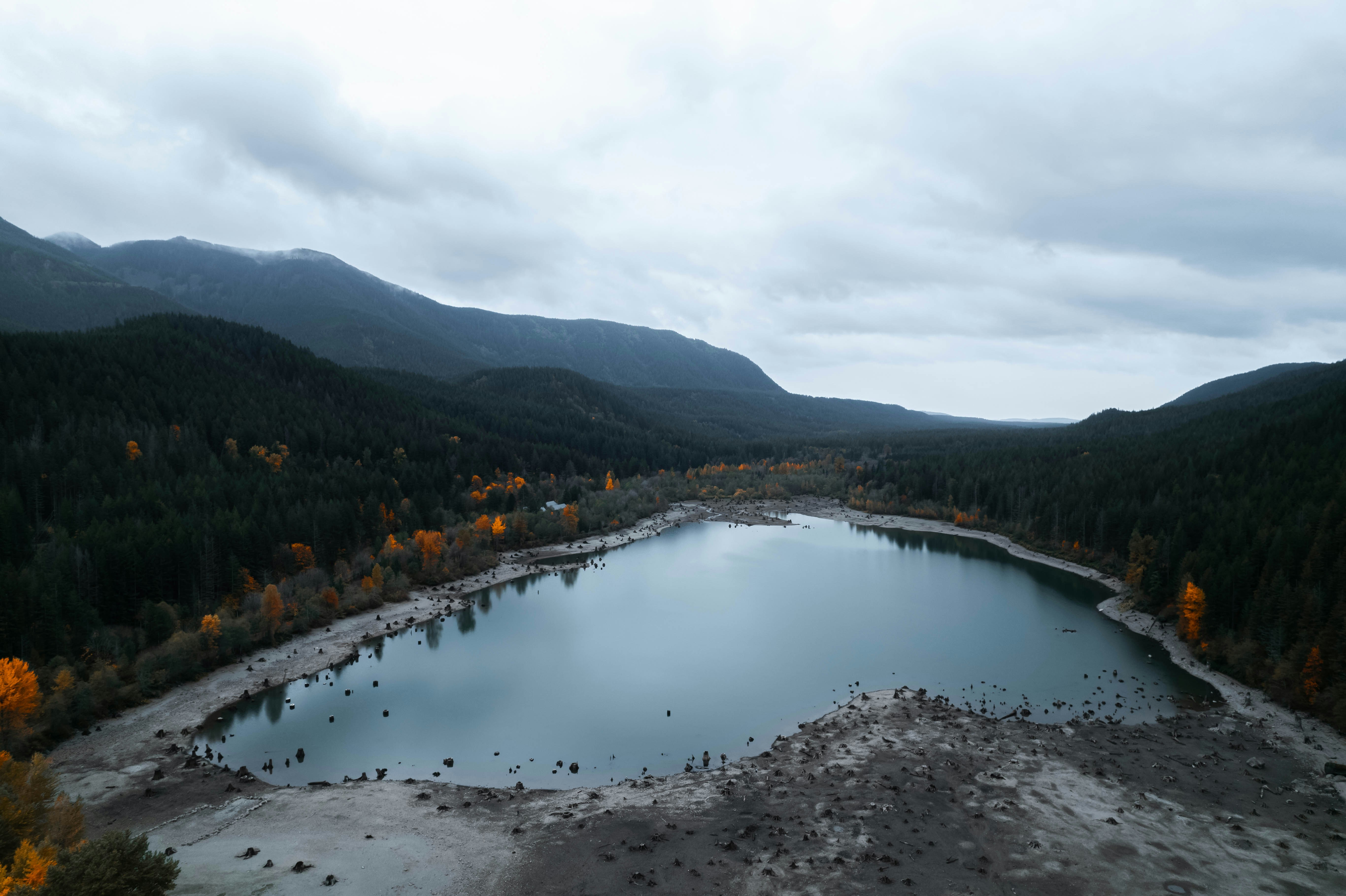Tranquil lake surrounded by dense forest, with autumn foliage reflecting on the water's surface. Overcast skies add a moody atmosphere.