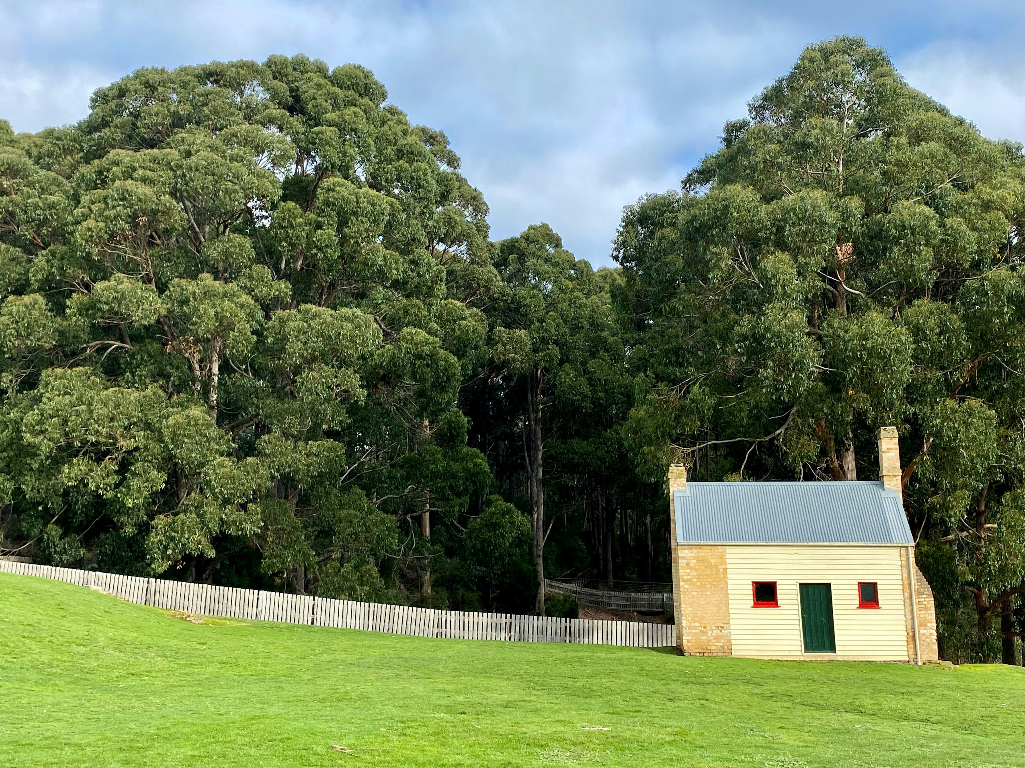 Small yellow cottage with a green door, nestled against tall, lush trees under a cloudy sky.