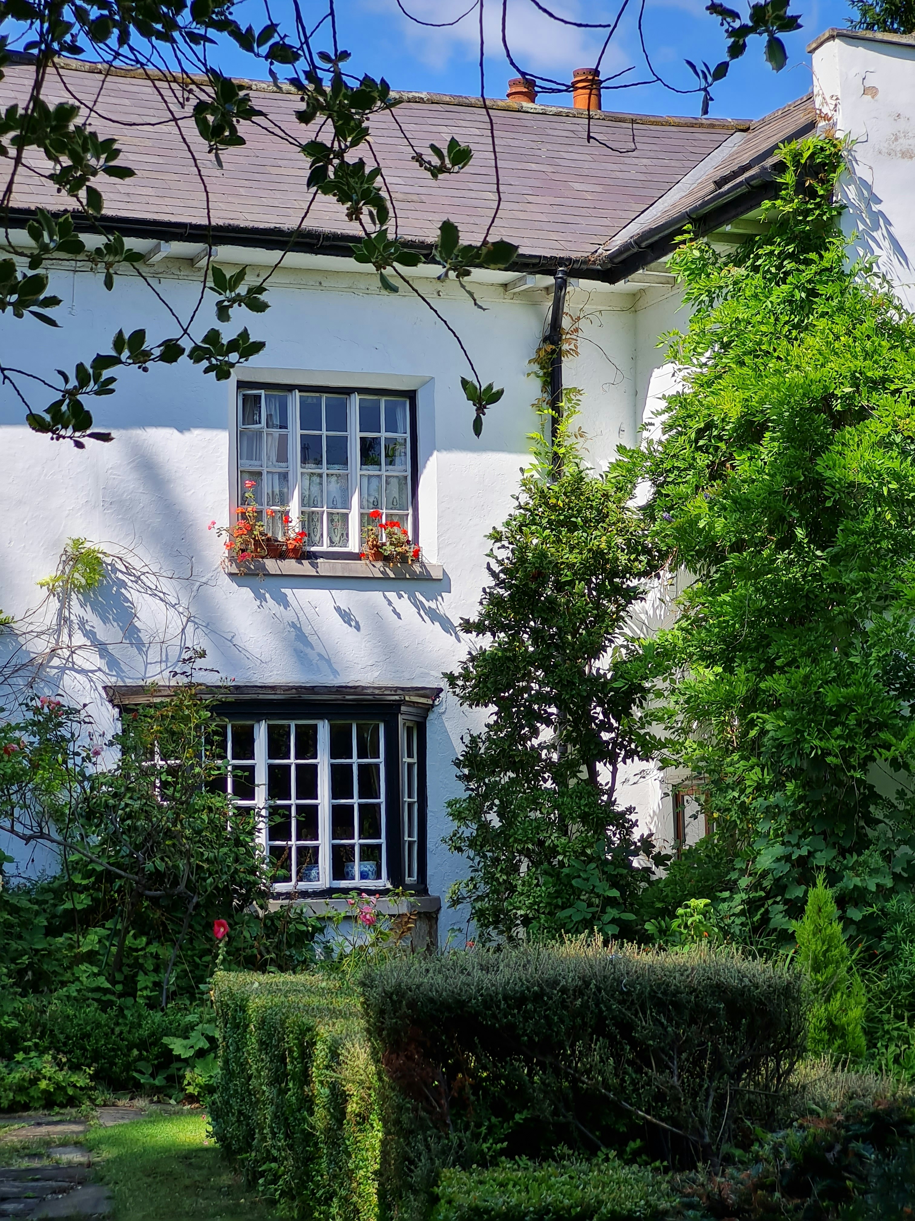 Photograph of a white two-story cottage with flower-box windows and a lush garden in front. The scene captures a sunlit domestic exterior with vibrant greenery.