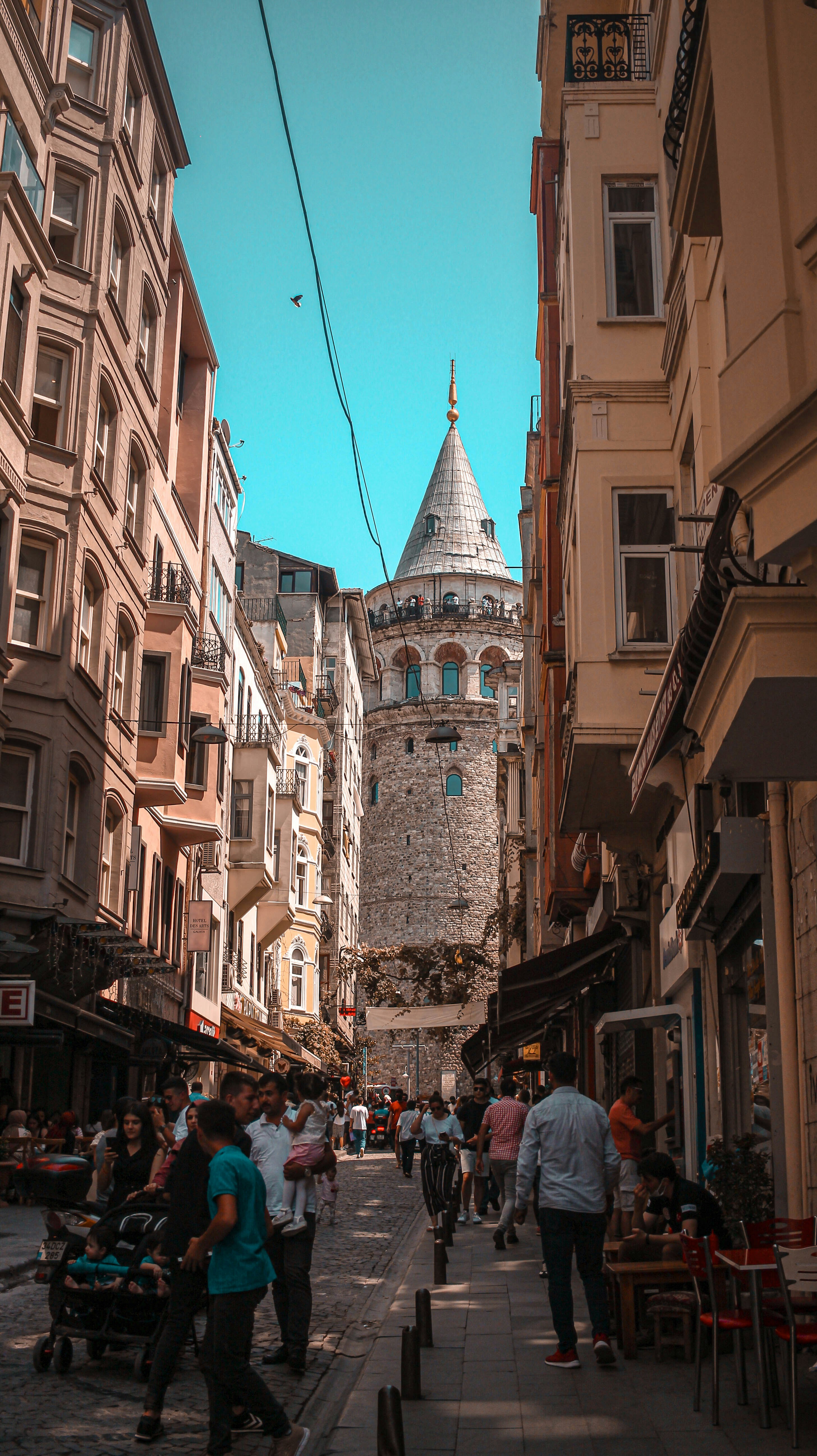 Galata Tower rises between narrow streets bustling with people under a clear blue sky.
