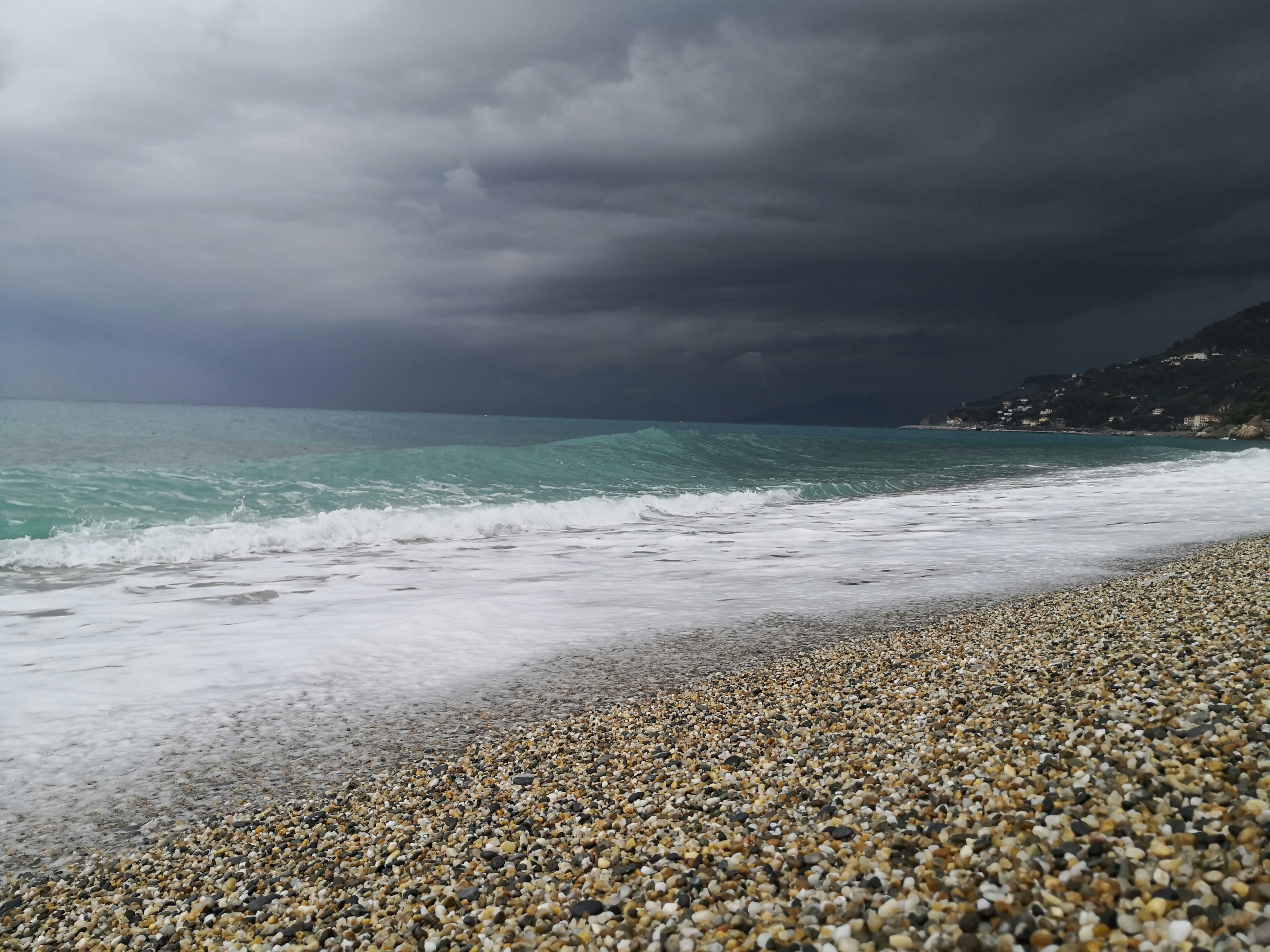Pebble beach stretches along turquoise water as dark storm clouds roll in and waves break on the shore.