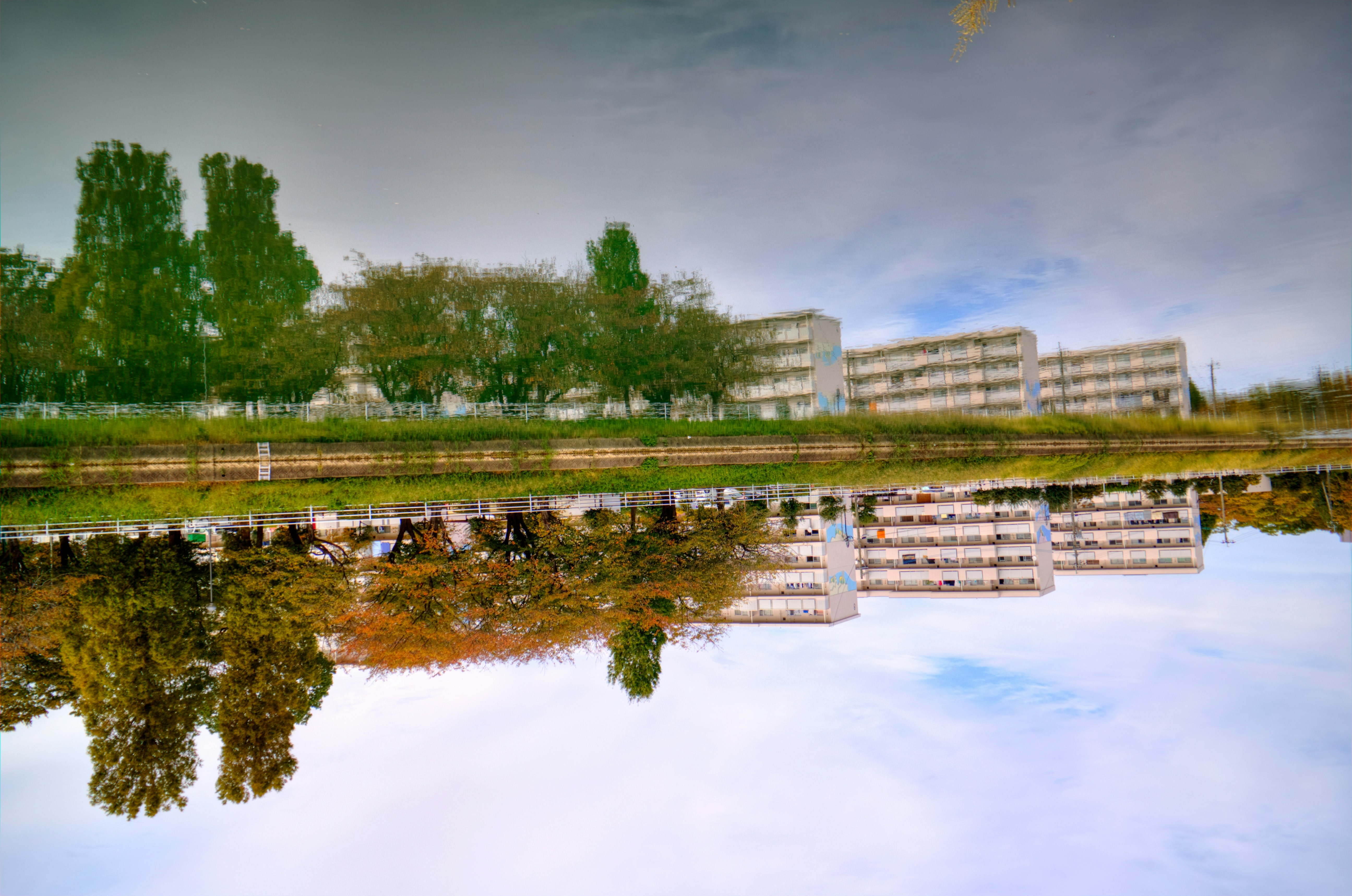 A tranquil scene showcasing the reflection of residential buildings and trees on a calm water surface. The image captures a harmonious blend of nature and urban architecture.