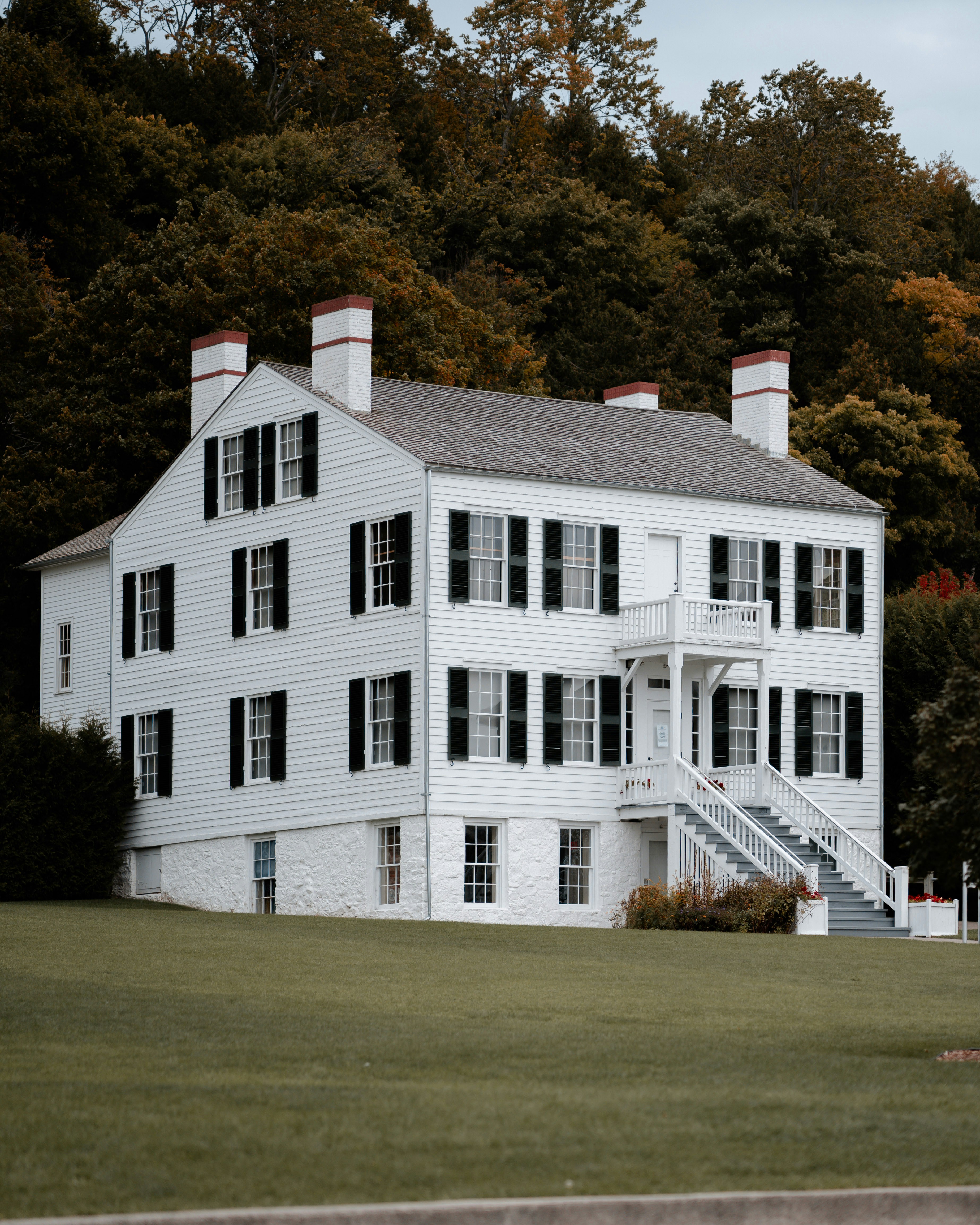 Classic white colonial house with black shutters and a welcoming porch, surrounded by lush greenery and autumn foliage.