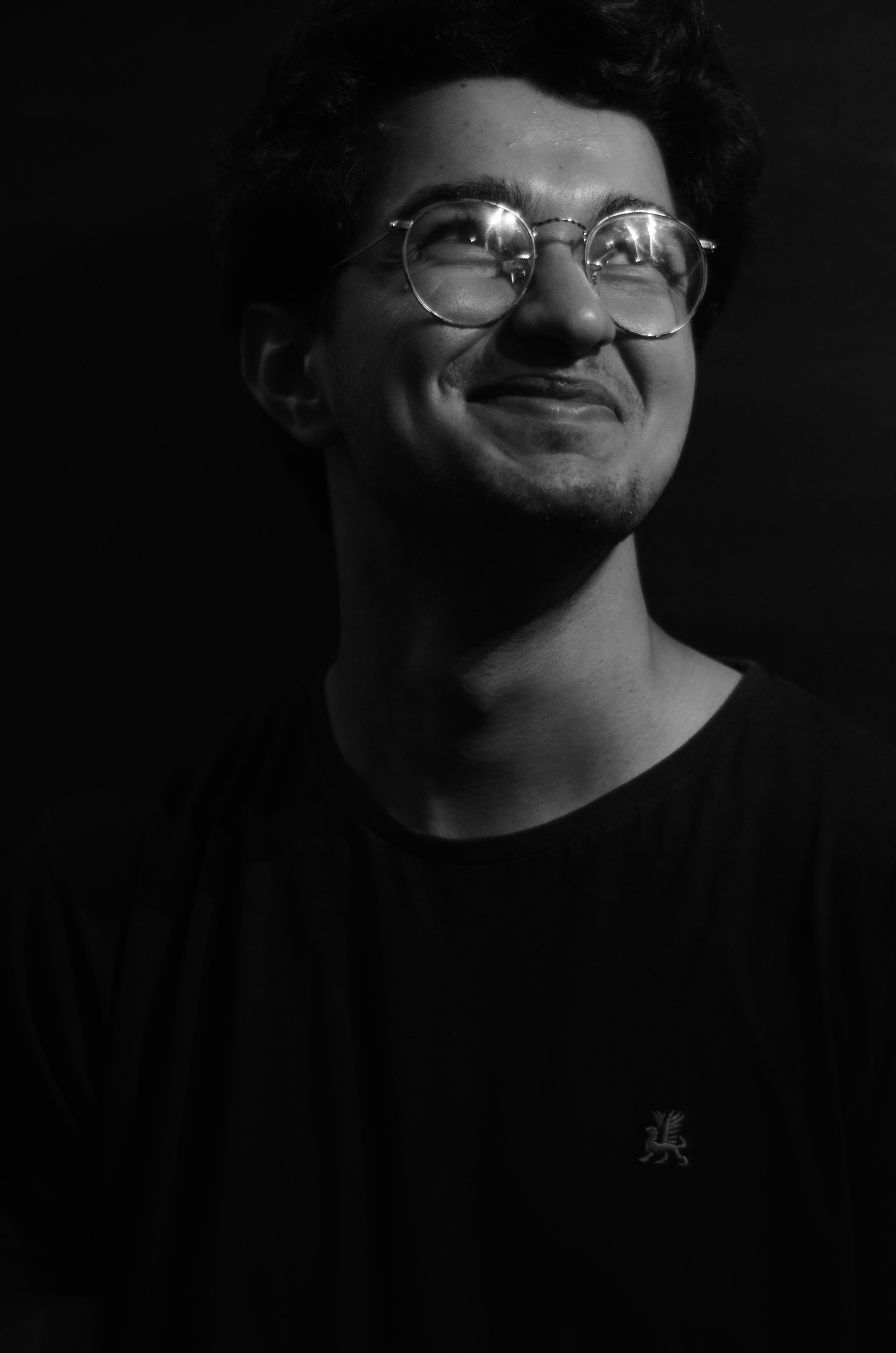 A young man with glasses smiles softly against a dark background, showcasing a moment of genuine happiness.