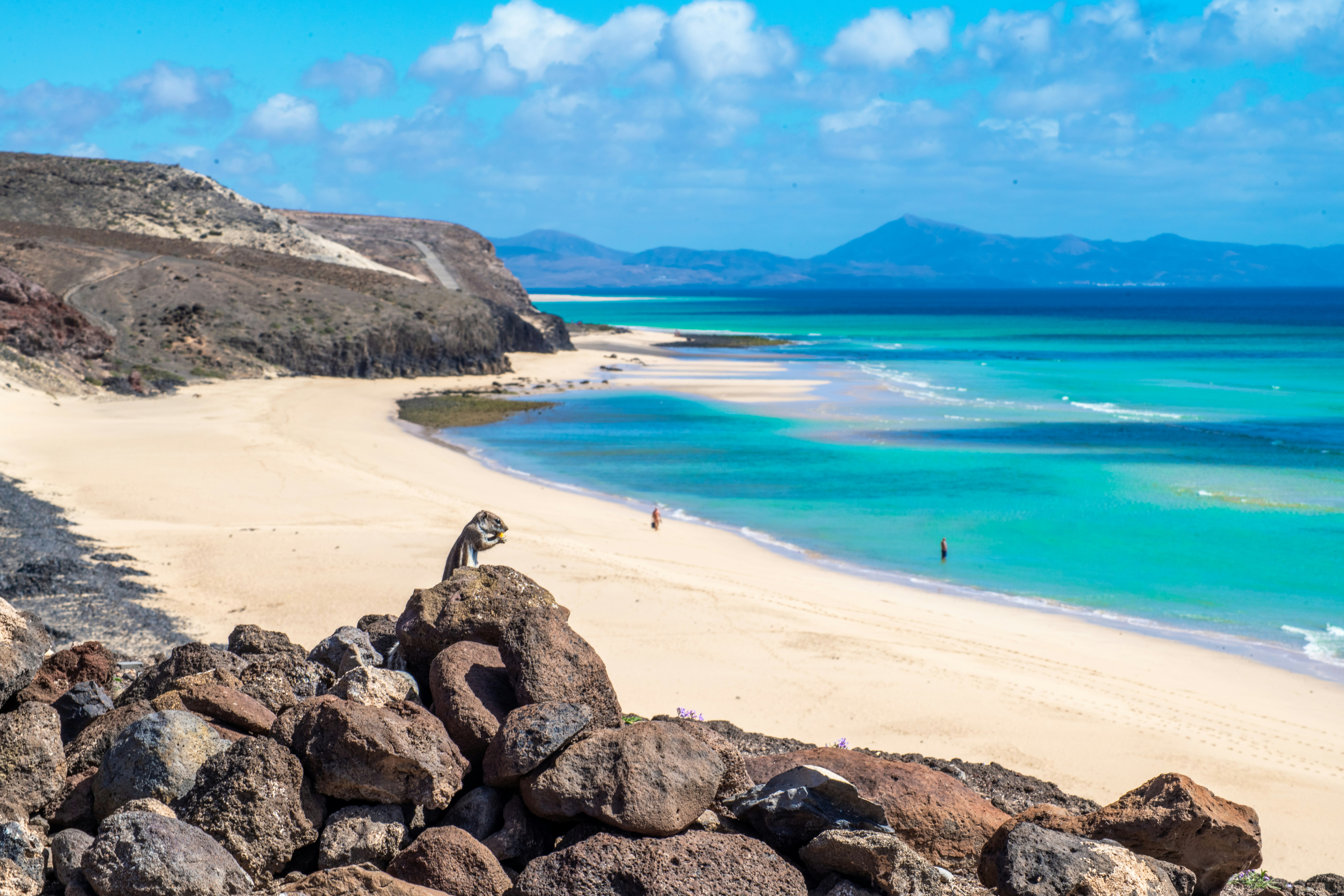 Tranquil beach scene featuring gentle waves lapping against a sandy shore, framed by rocky formations and distant mountains.