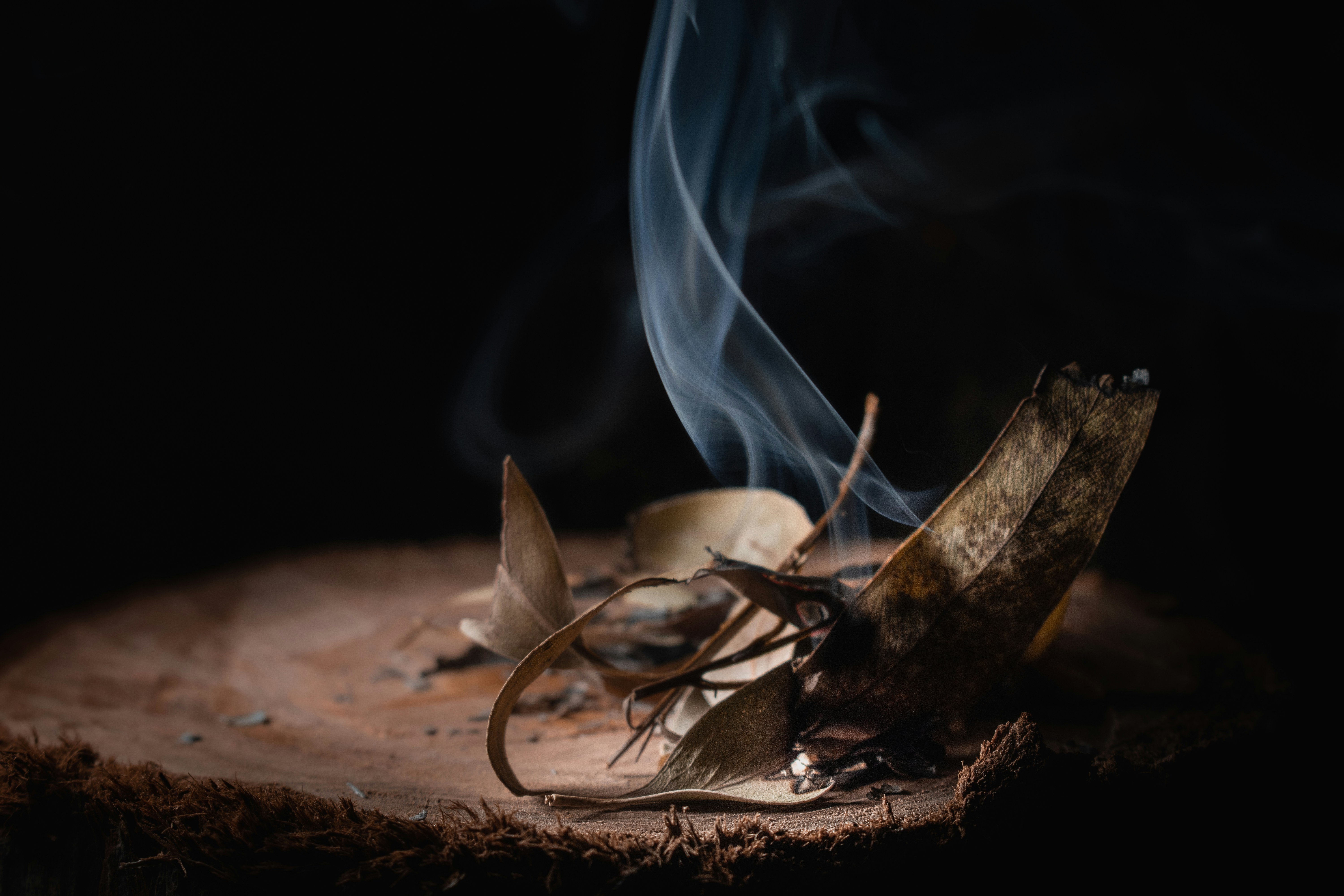 Dried leaves smolder on a wooden surface, releasing delicate wisps of smoke against a dark backdrop.