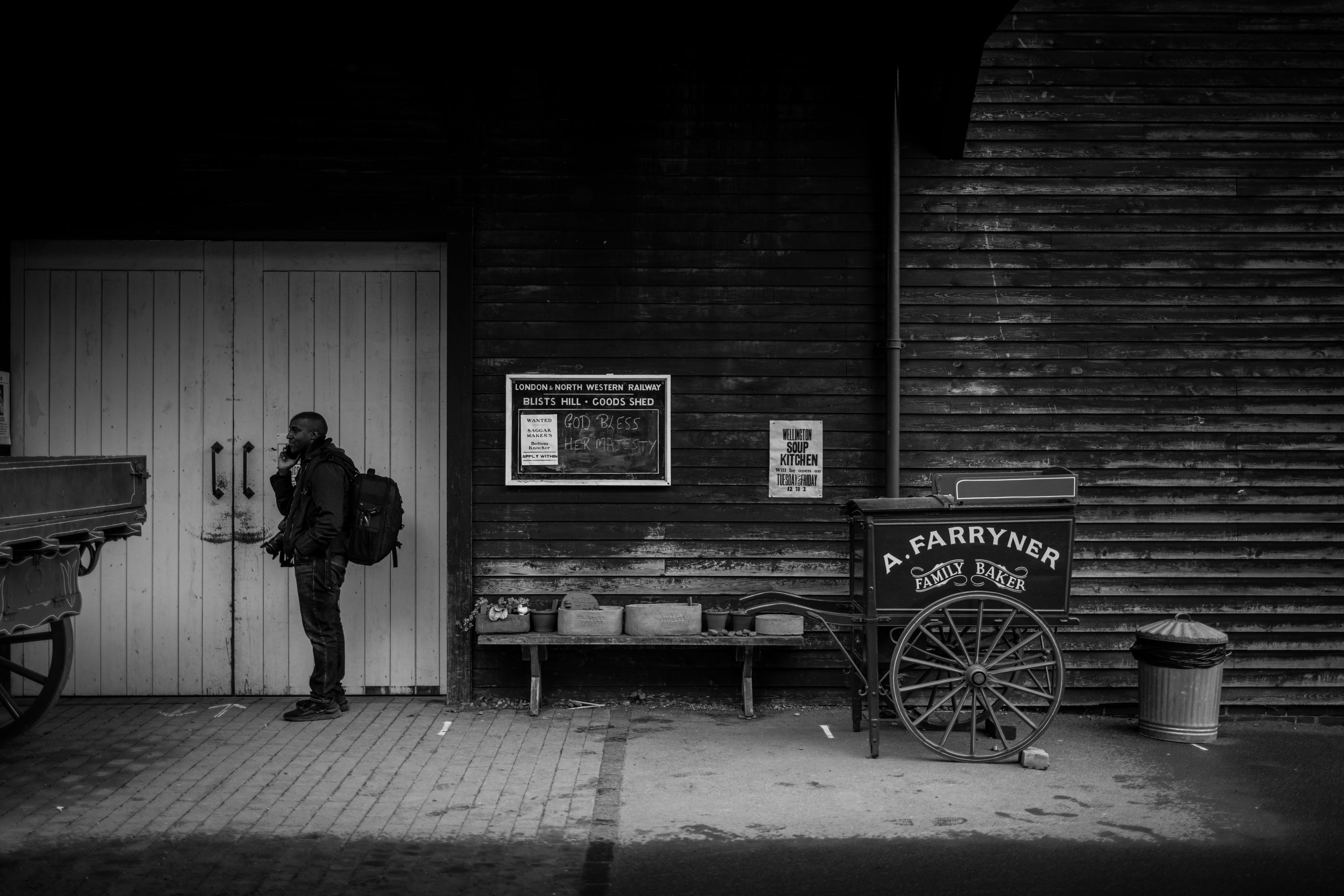 Person with a backpack stands near a closed barn door beside a vintage cart in a dimly lit alley.