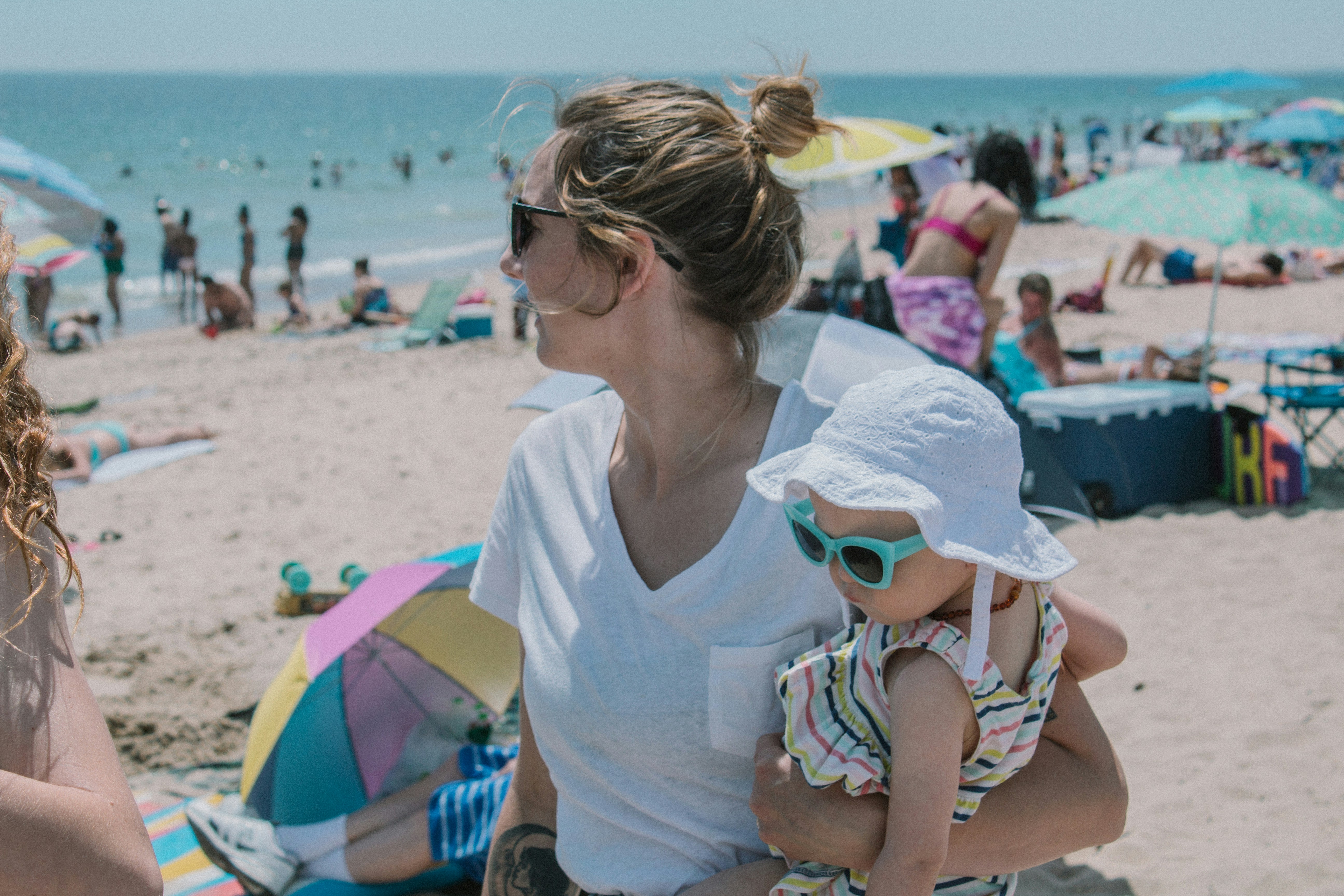 Woman holding a child in sunglasses and a hat on a lively beach with colorful umbrellas.