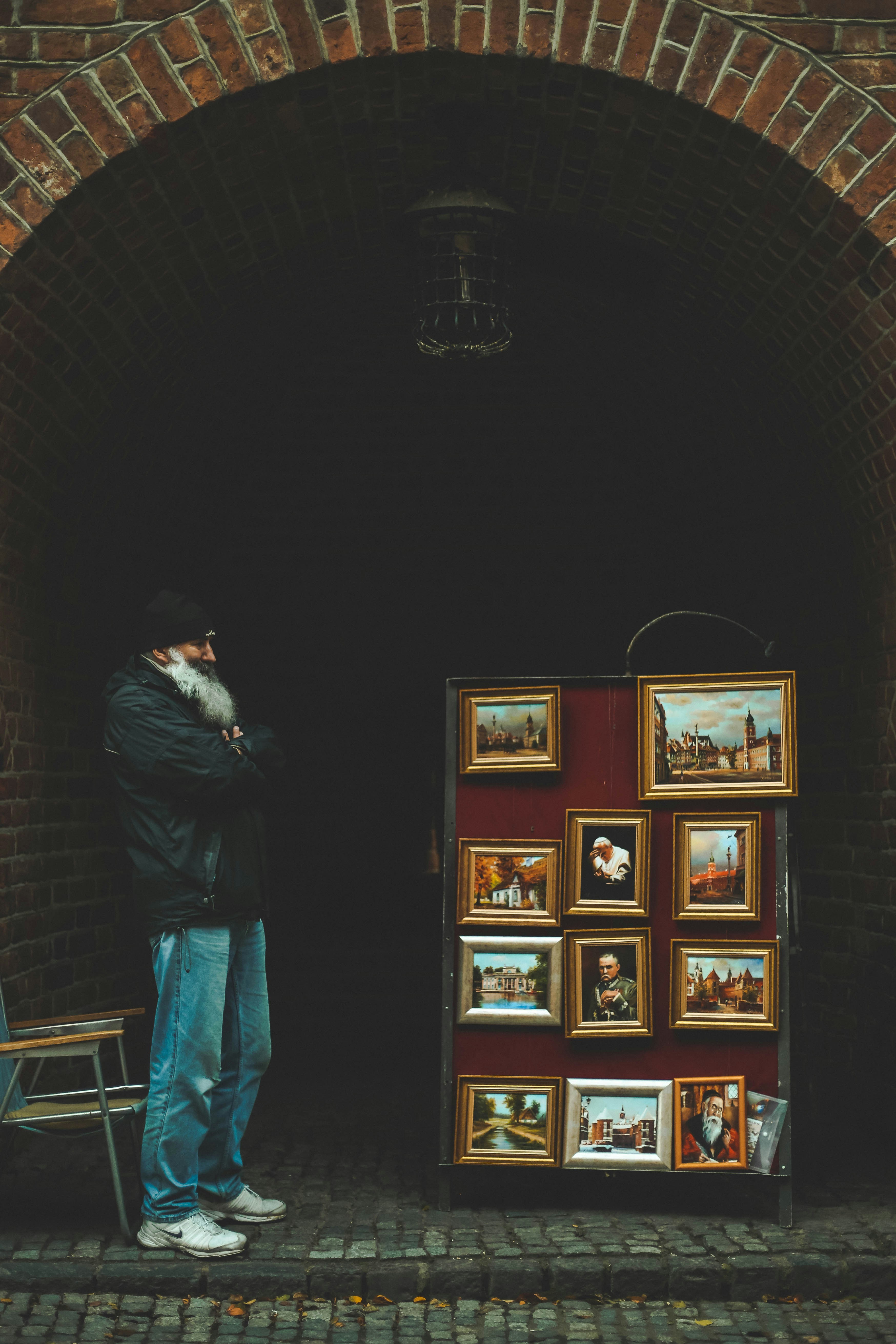 An elderly man stands beside a display of framed artworks in a dimly lit archway, showcasing a blend of historical and contemporary scenes.