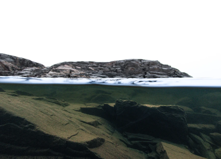 Half-above, half-below water view of a rocky shoreline, with bright sky above and submerged rocks visible in the greenish water below. The surface line cuts horizontally across the frame.
