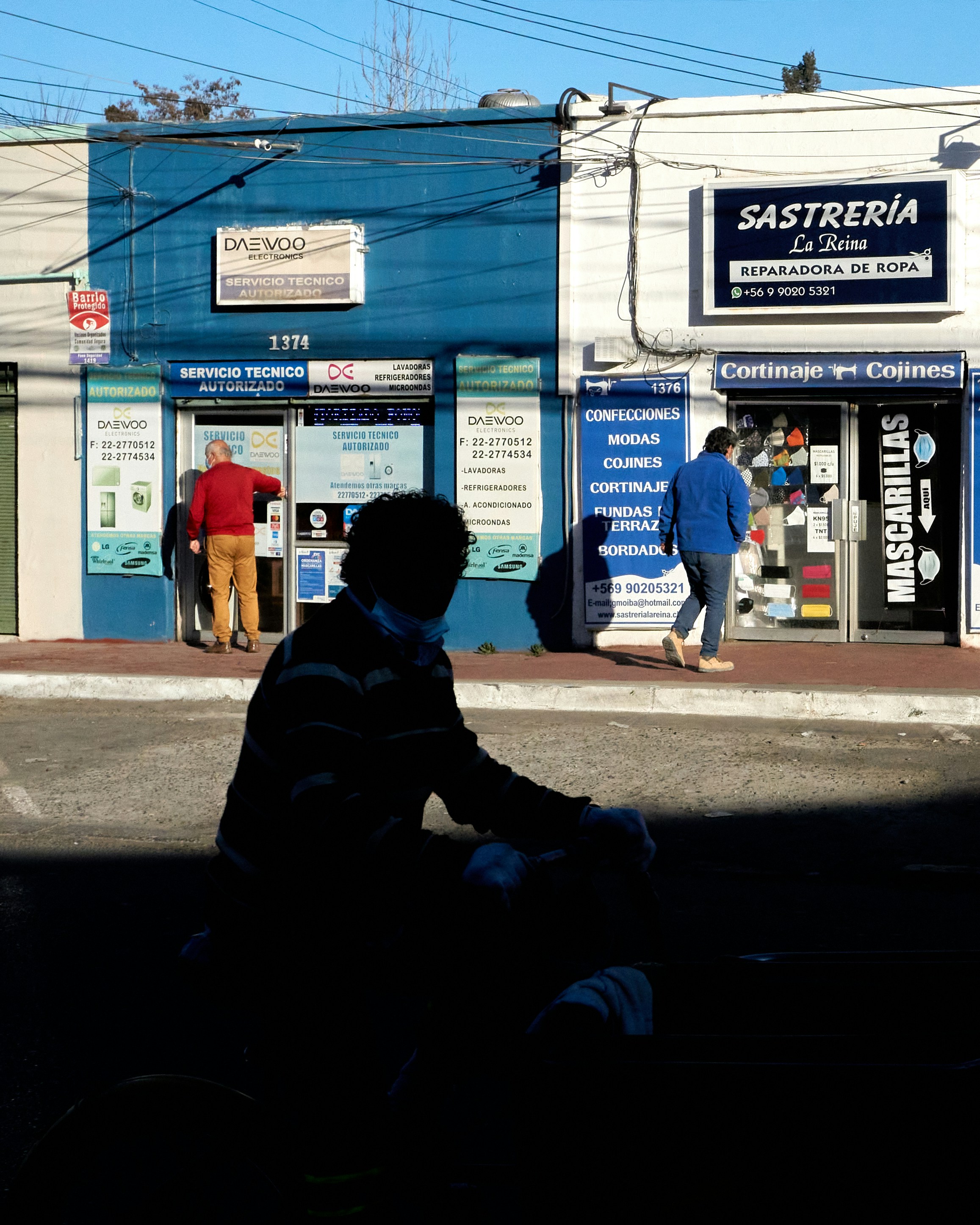 Local tailor shop and technical service storefronts bustling with activity. A silhouette of a person in the foreground adds depth to the scene.