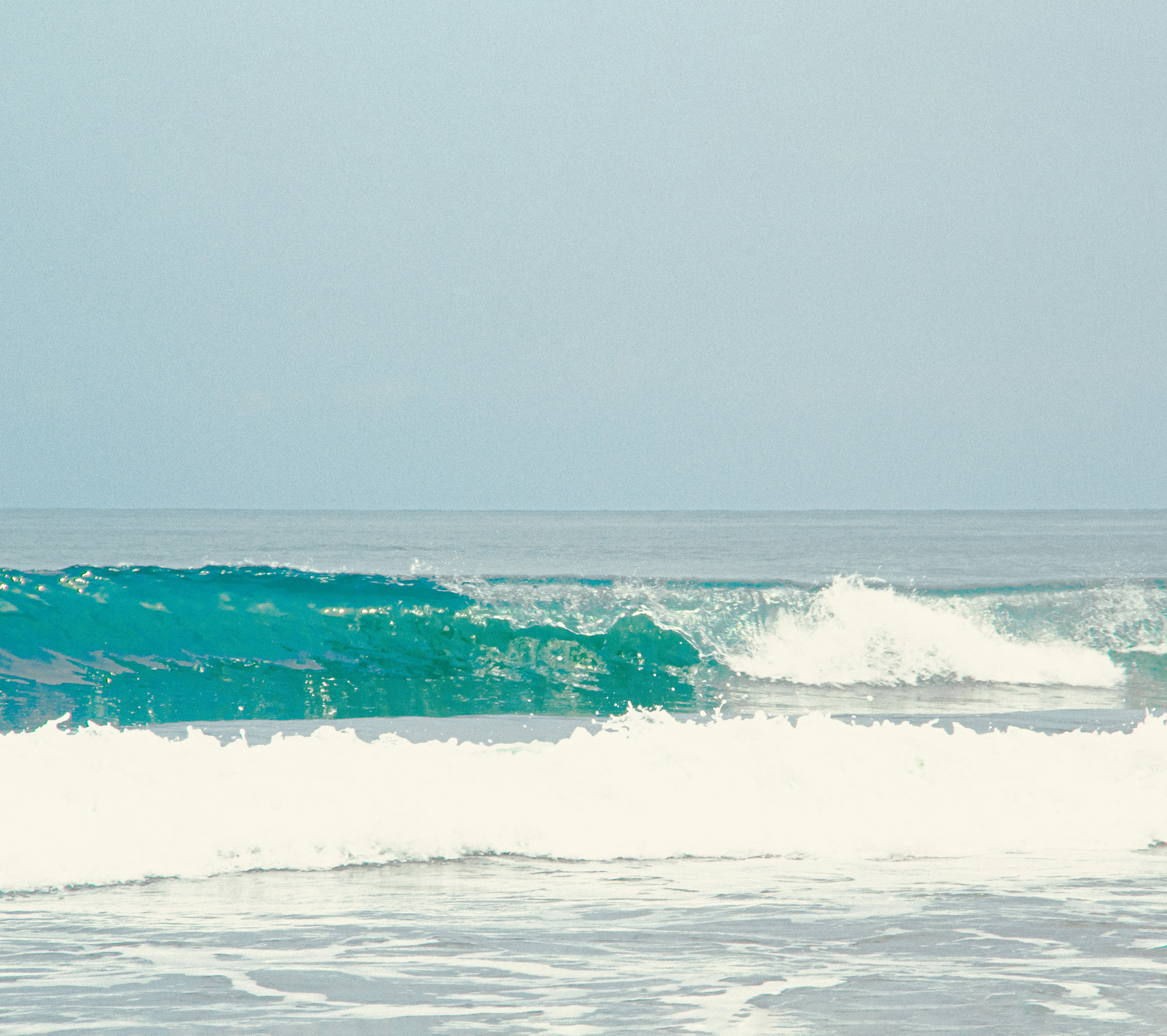 Vibrant turquoise waves cresting against a serene shoreline under a soft, overcast sky.