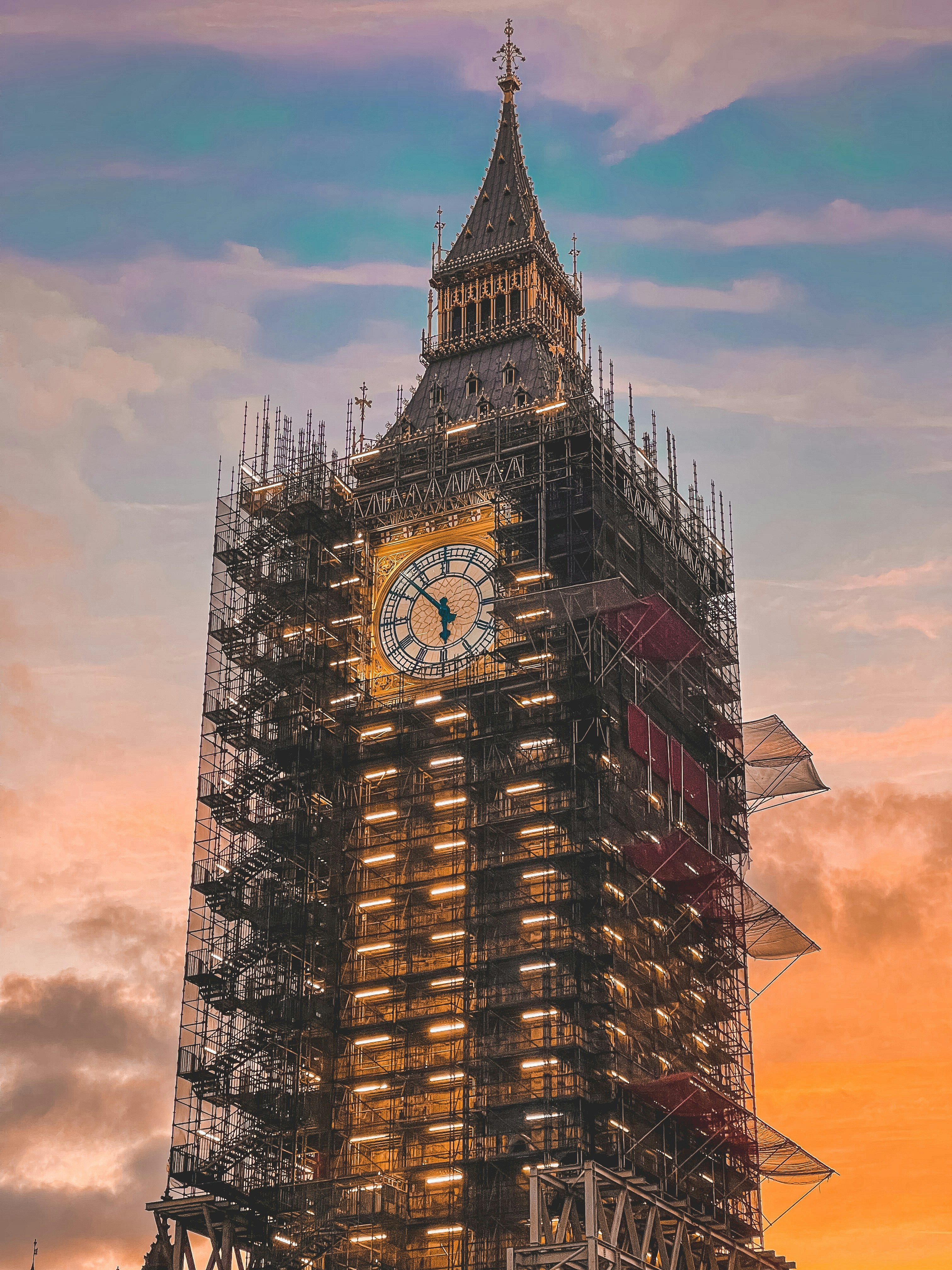 Big Ben enveloped in scaffolding during restoration, showcasing its iconic clock face against a colorful sunset.