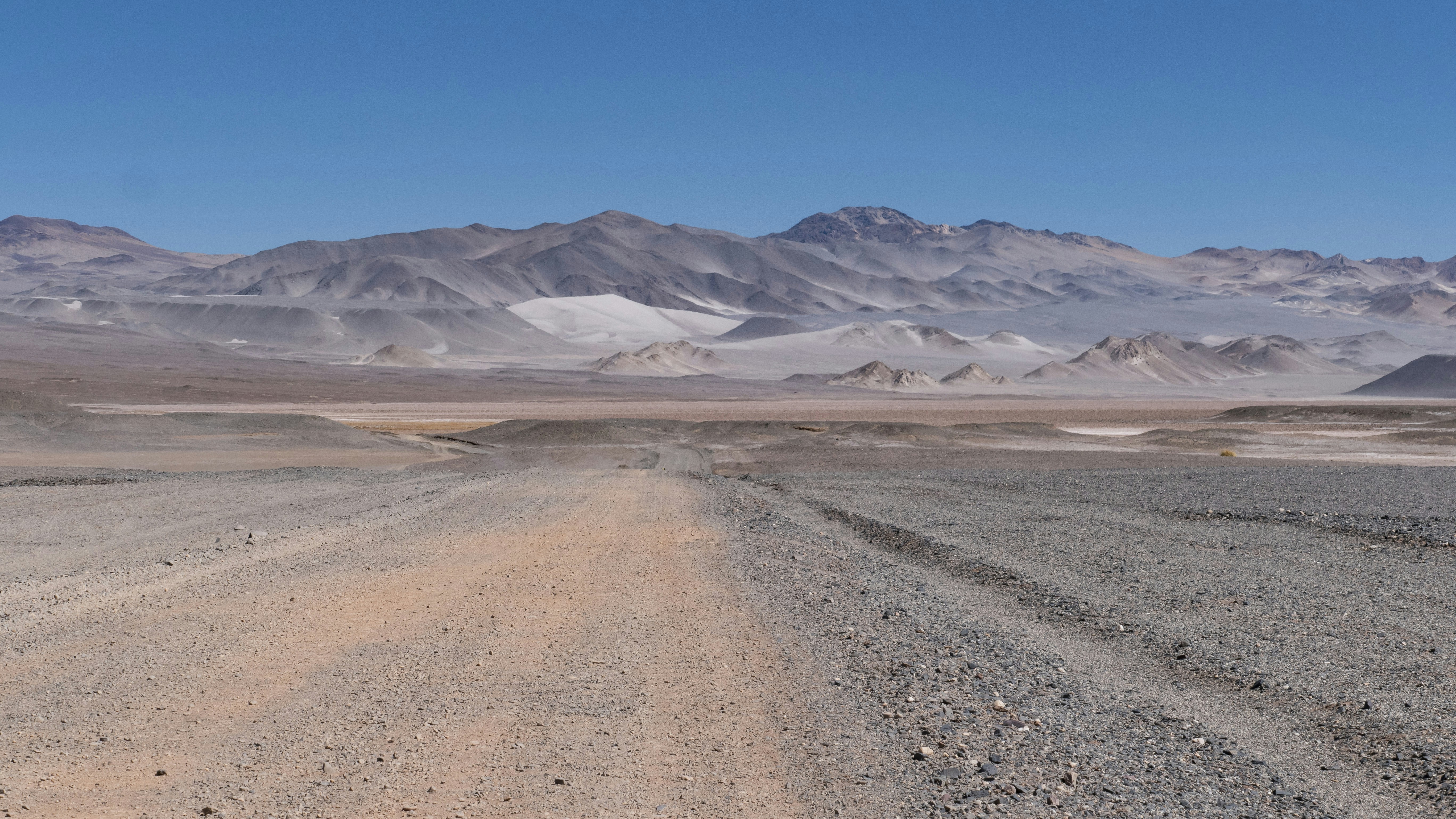 Rugged dirt road winding through a vast, barren landscape with distant mountains under a clear blue sky.