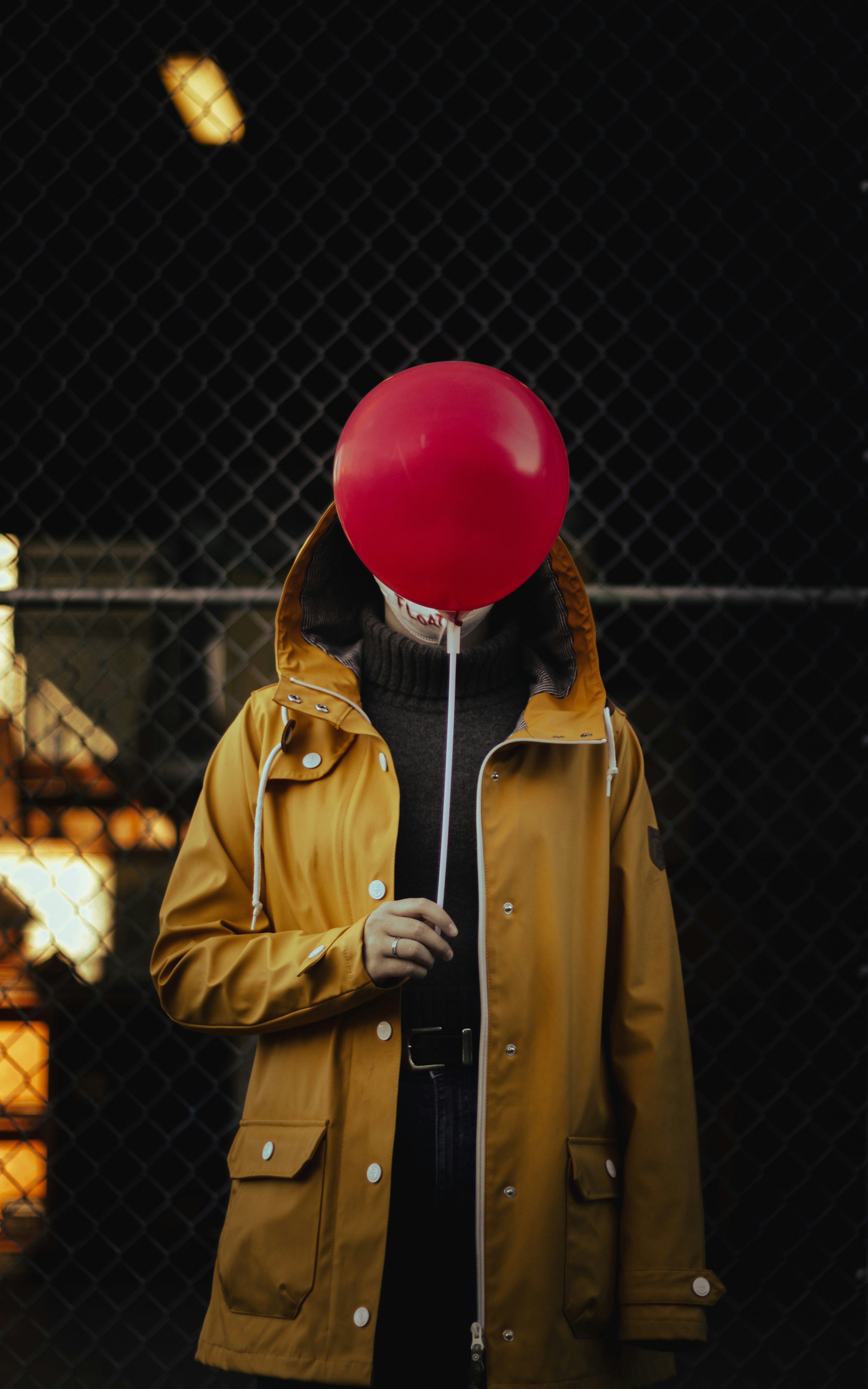 Individual in a yellow raincoat holding a vibrant red balloon against a dark, textured background. The scene evokes a sense of mystery and intrigue.