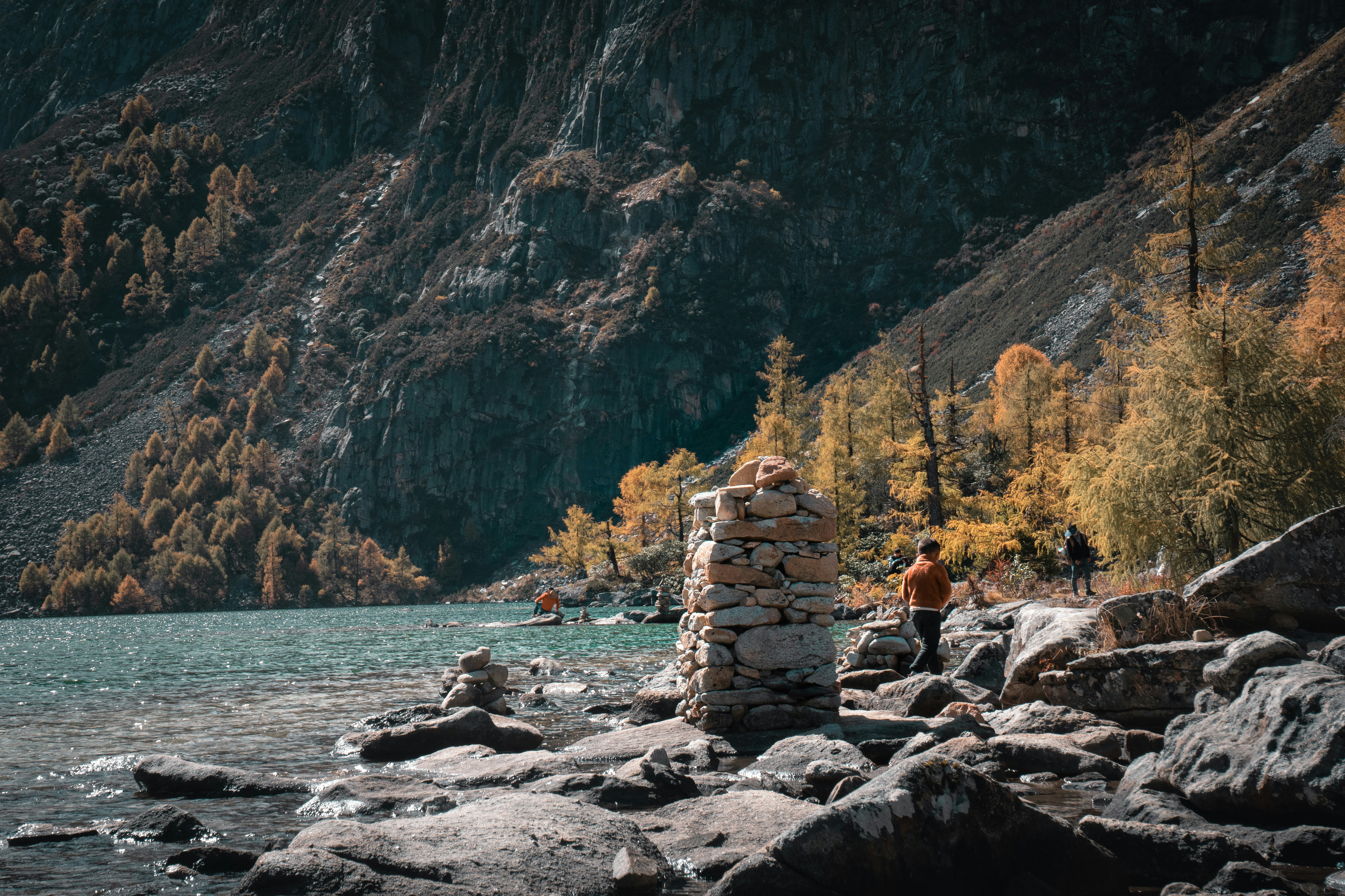 A serene lakeside scene featuring a stone cairn surrounded by autumn foliage and individuals enjoying the tranquil environment.