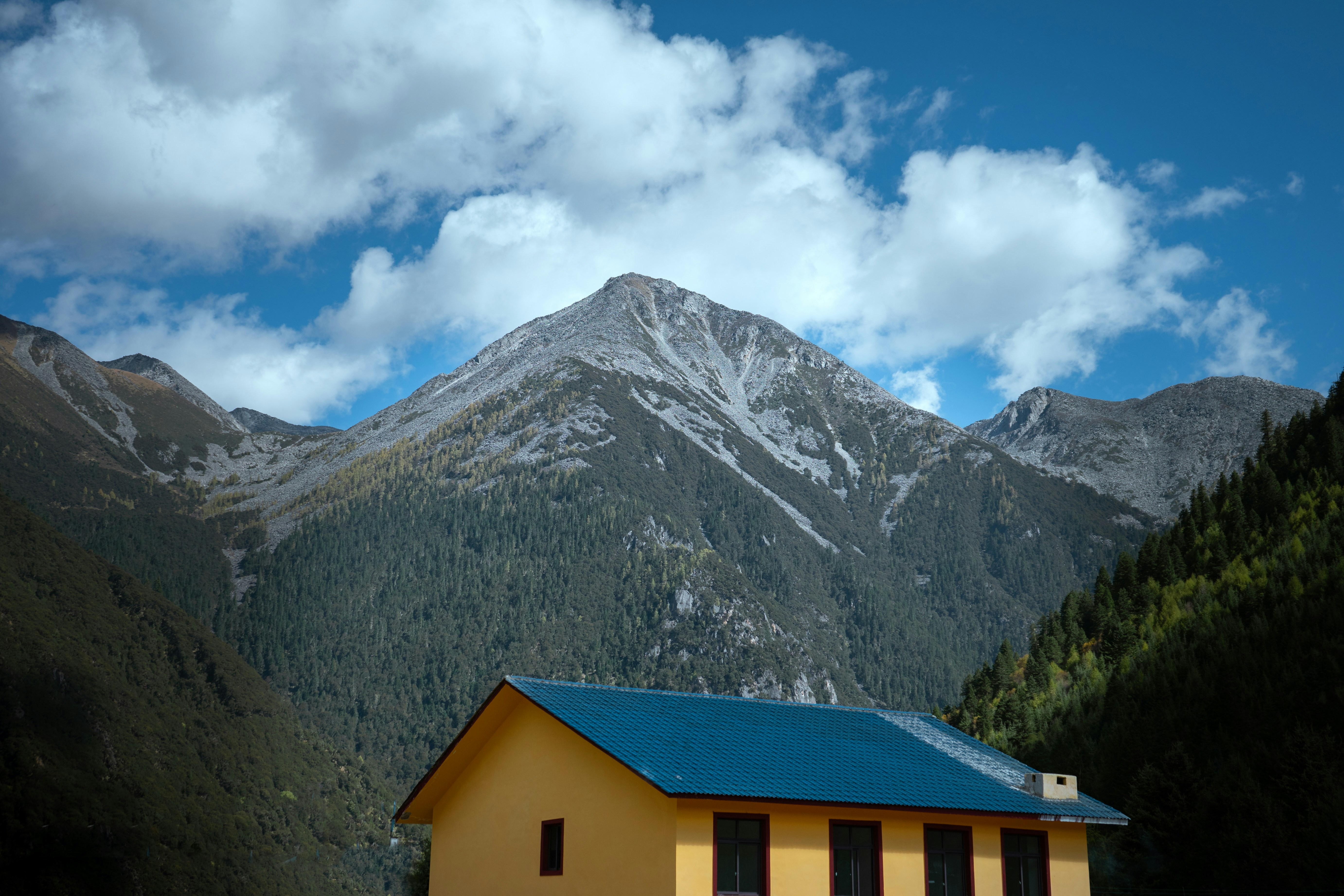 A vibrant yellow house nestled at the foot of majestic mountains under a bright blue sky with scattered clouds.