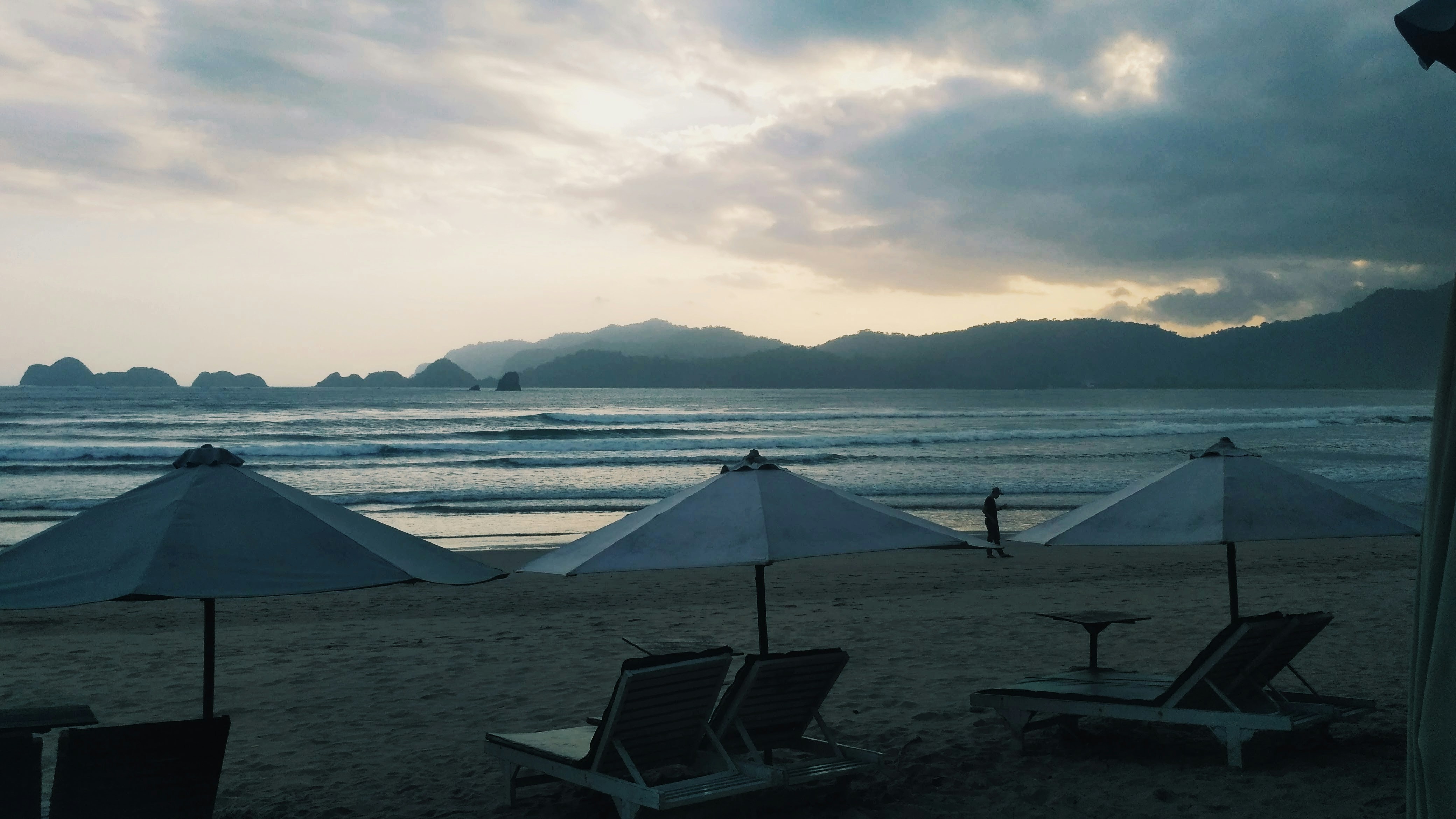 Beach scene at dusk with white umbrellas, lounge chairs, and a solitary figure along the waterline beneath a cloudy sky.
