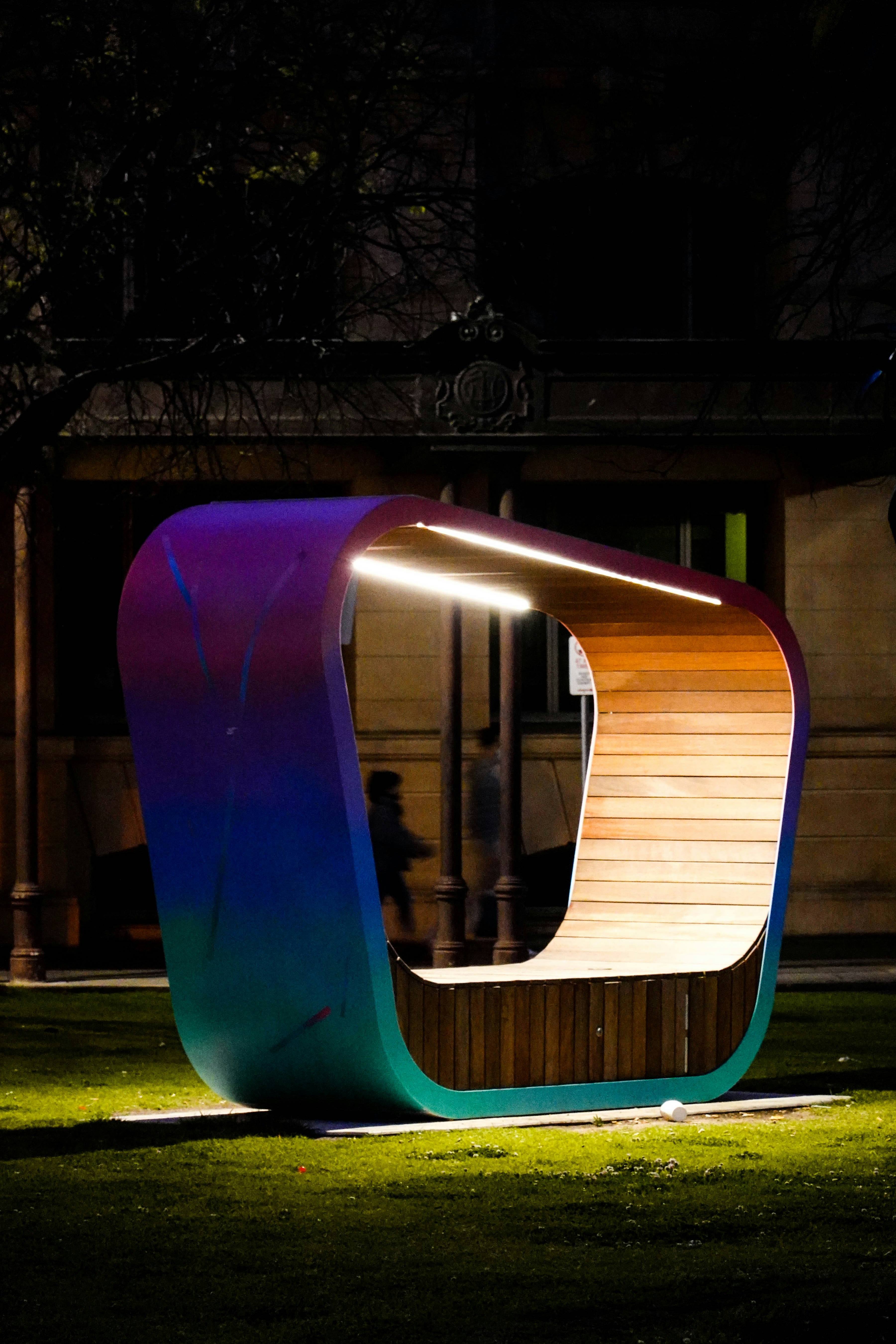 A vibrant, modern seating structure with a wooden interior and colorful exterior, illuminated at night, creating a striking contrast against the dark surroundings.