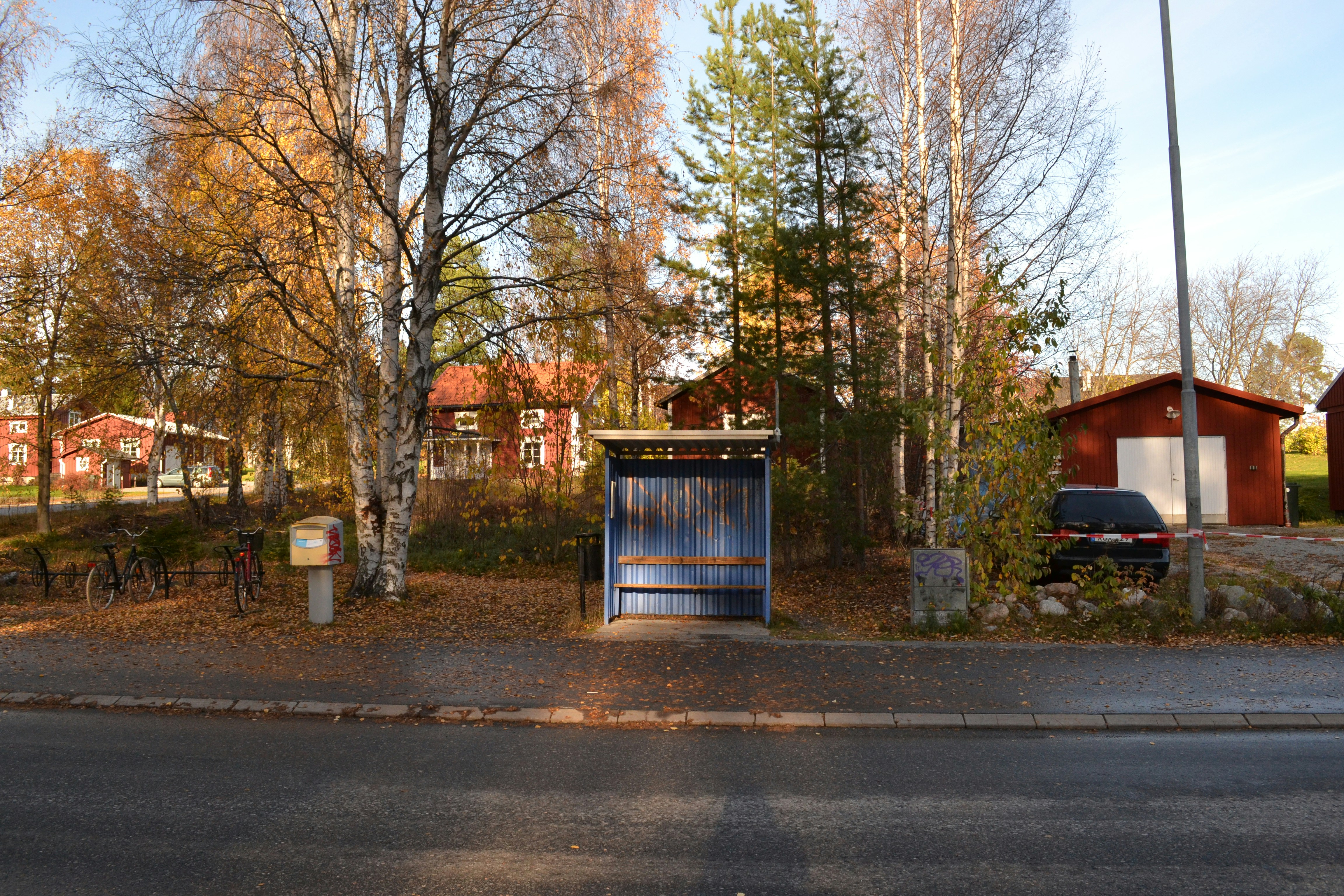 A tranquil bus stop nestled among autumn foliage, with red wooden houses in the background and bicycles parked nearby.