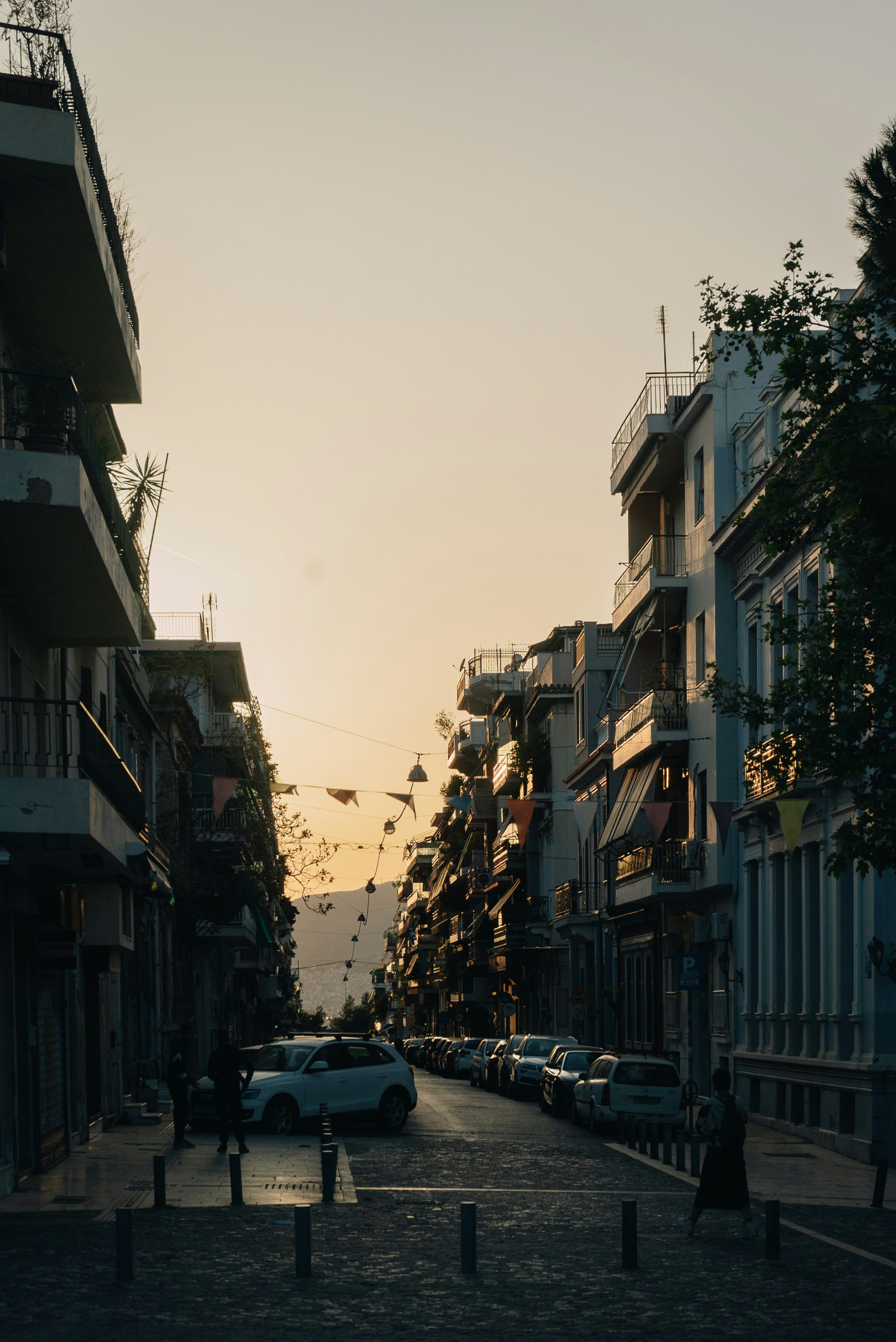 A serene street scene at dusk, framed by buildings and adorned with colorful flags, leading towards a soft sunset glow.
