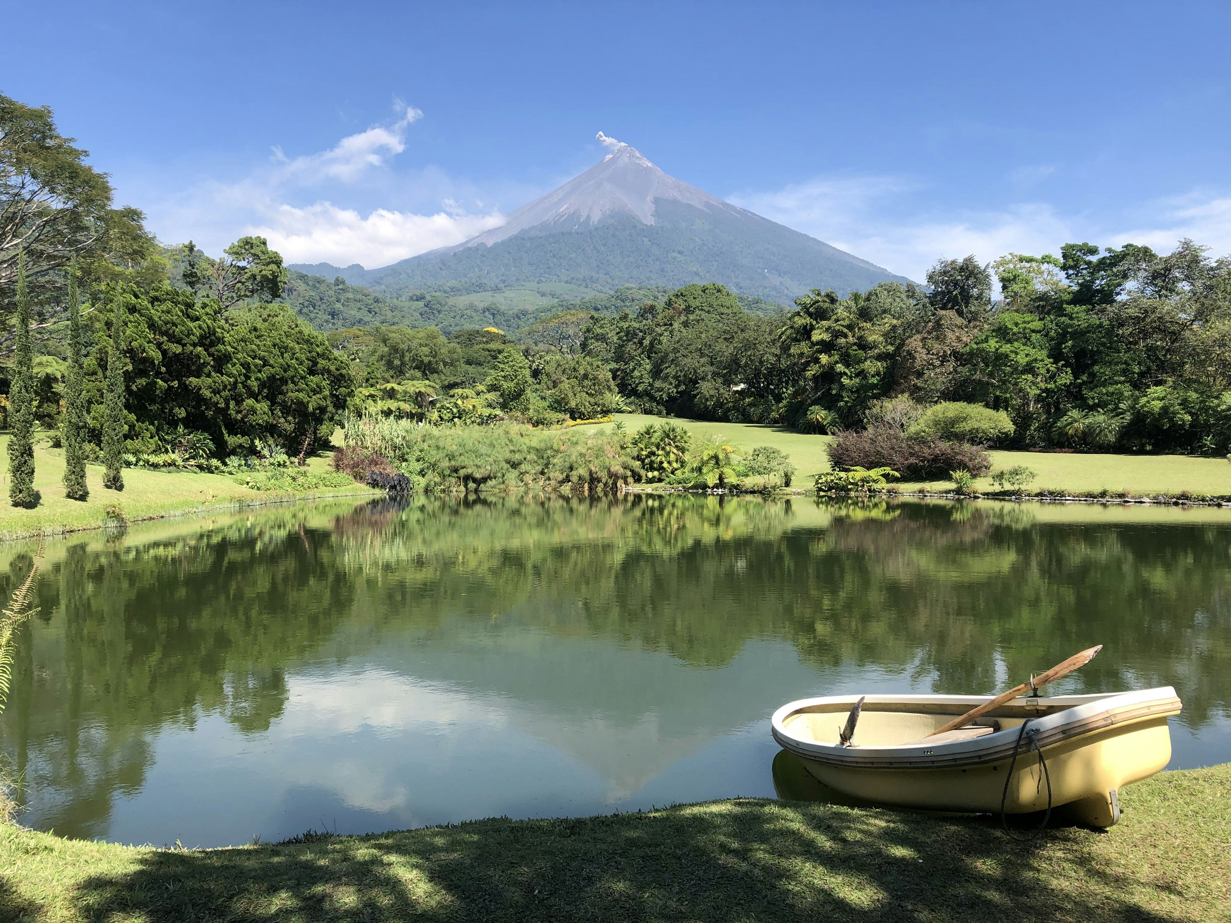A tranquil scene featuring a yellow boat resting by a reflective lake, framed by lush greenery and a majestic volcano in the background.