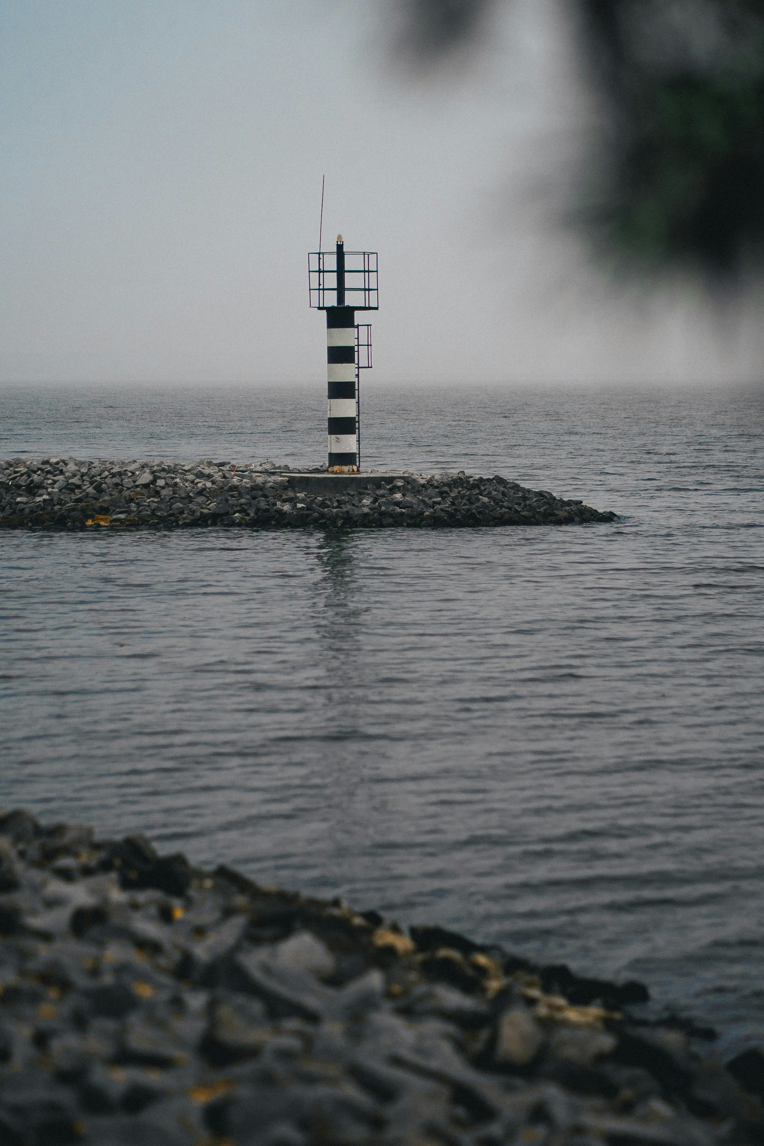 A solitary lighthouse stands on a rocky outcrop, surrounded by calm waters under a misty sky. The black and white stripes of the tower contrast with the serene landscape.