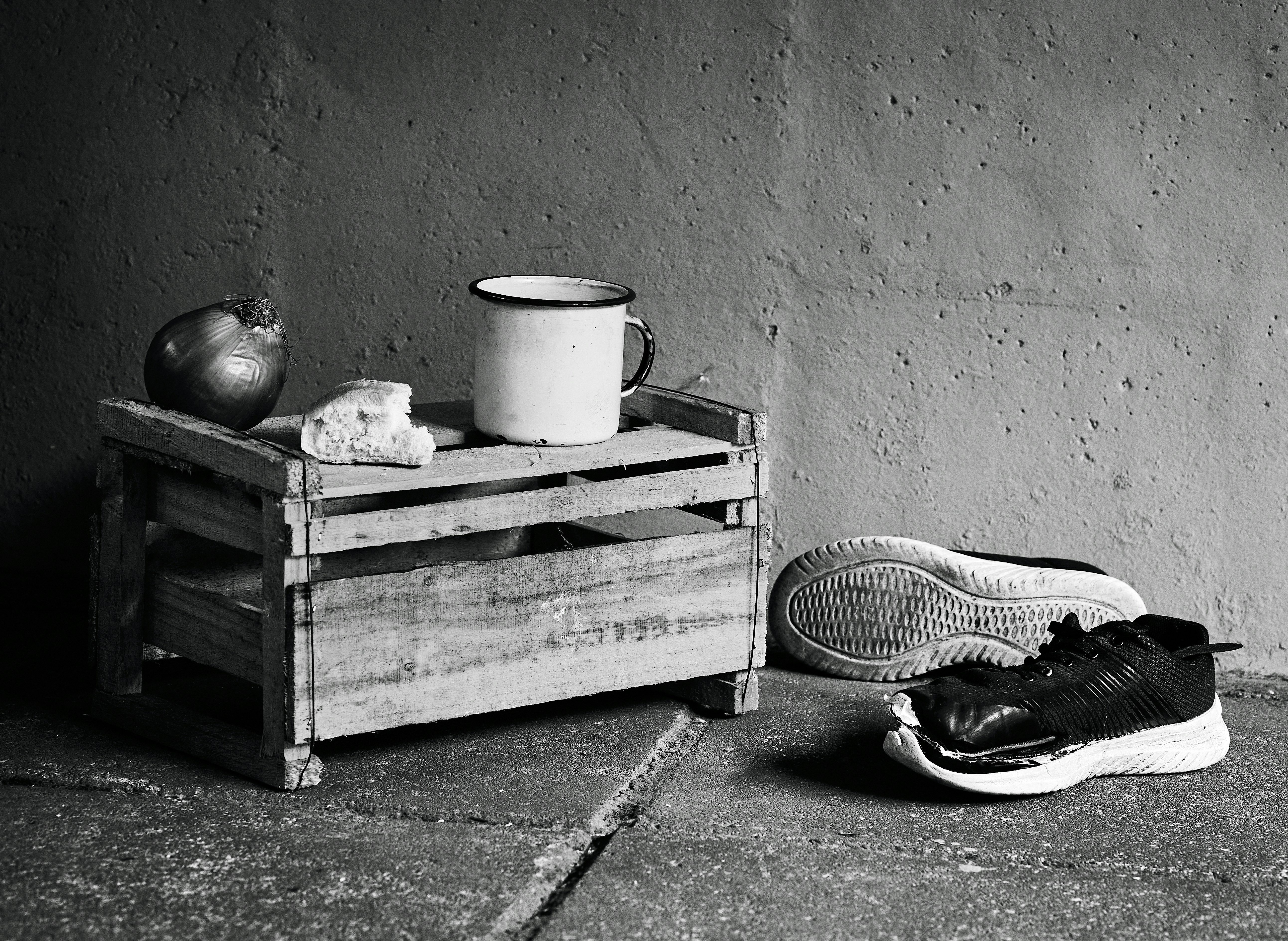Black and white still life with a wooden crate, an onion, a mug, a piece of bread, and worn sneakers on a textured floor.