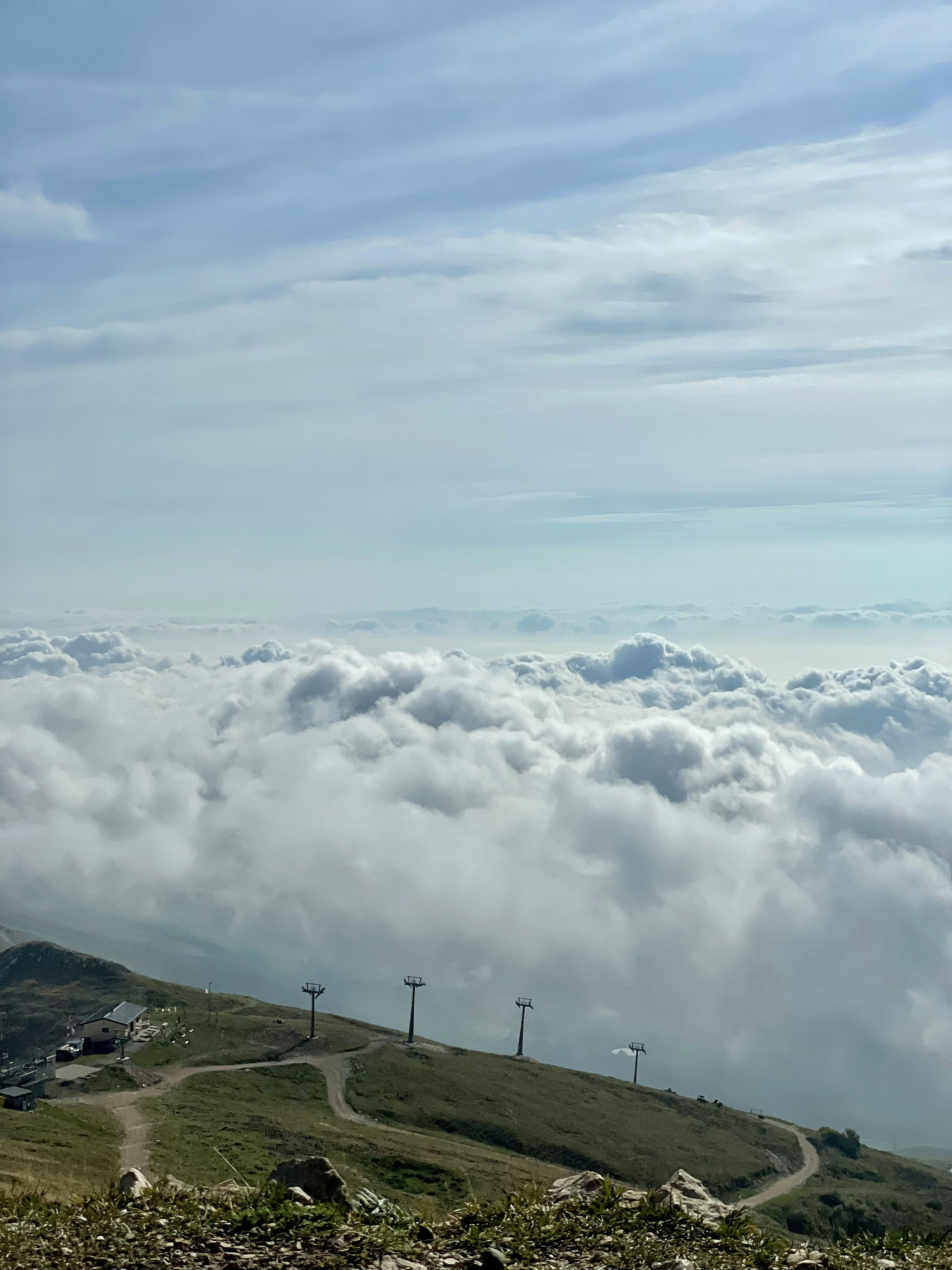 A panoramic view from a mountain peak, showcasing rolling clouds and a winding path leading to a ski lift. The scene captures the tranquility of high-altitude landscapes.