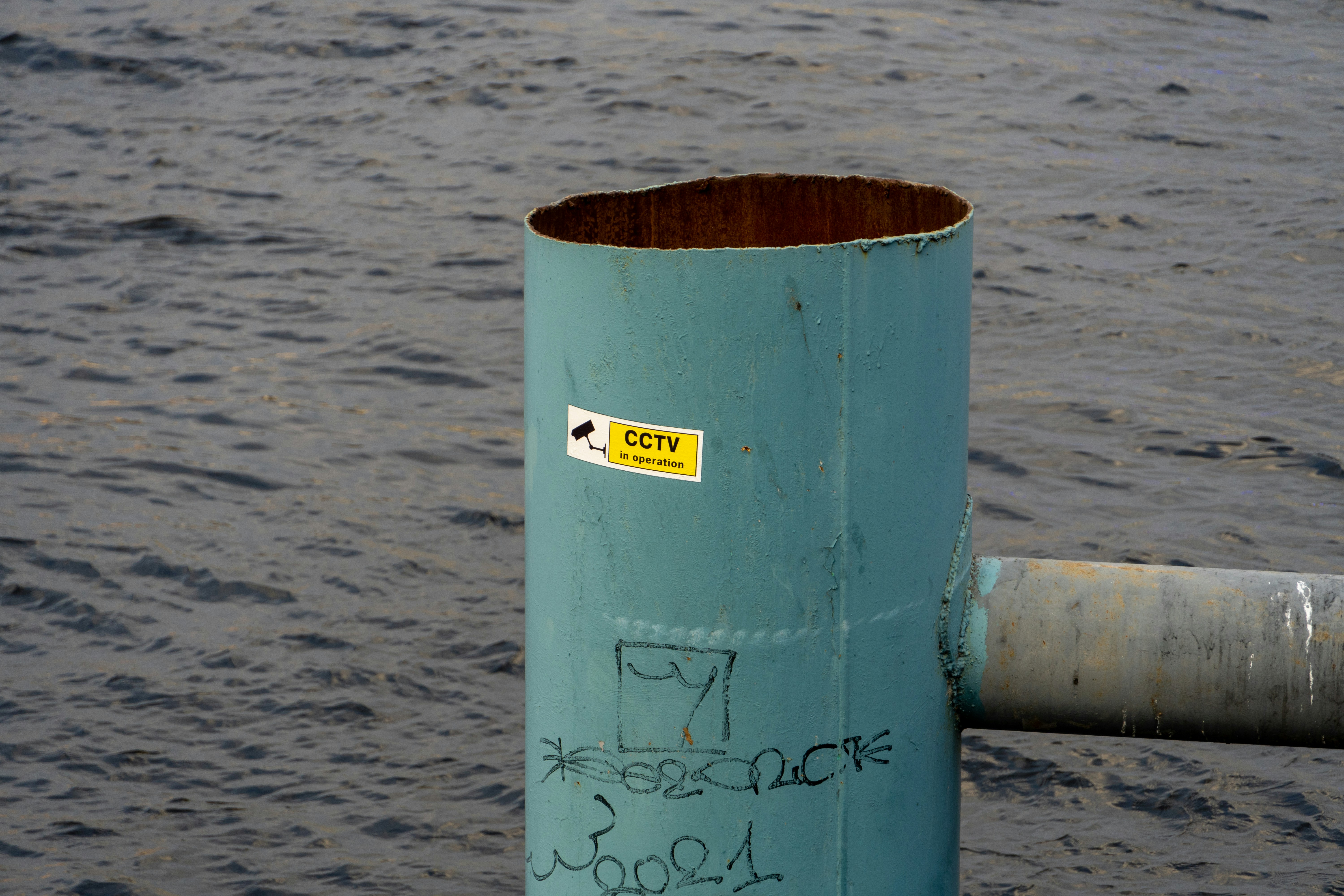 CCTV Label on a Weathered Blue Pipe Over Quiet Harbor Ripples