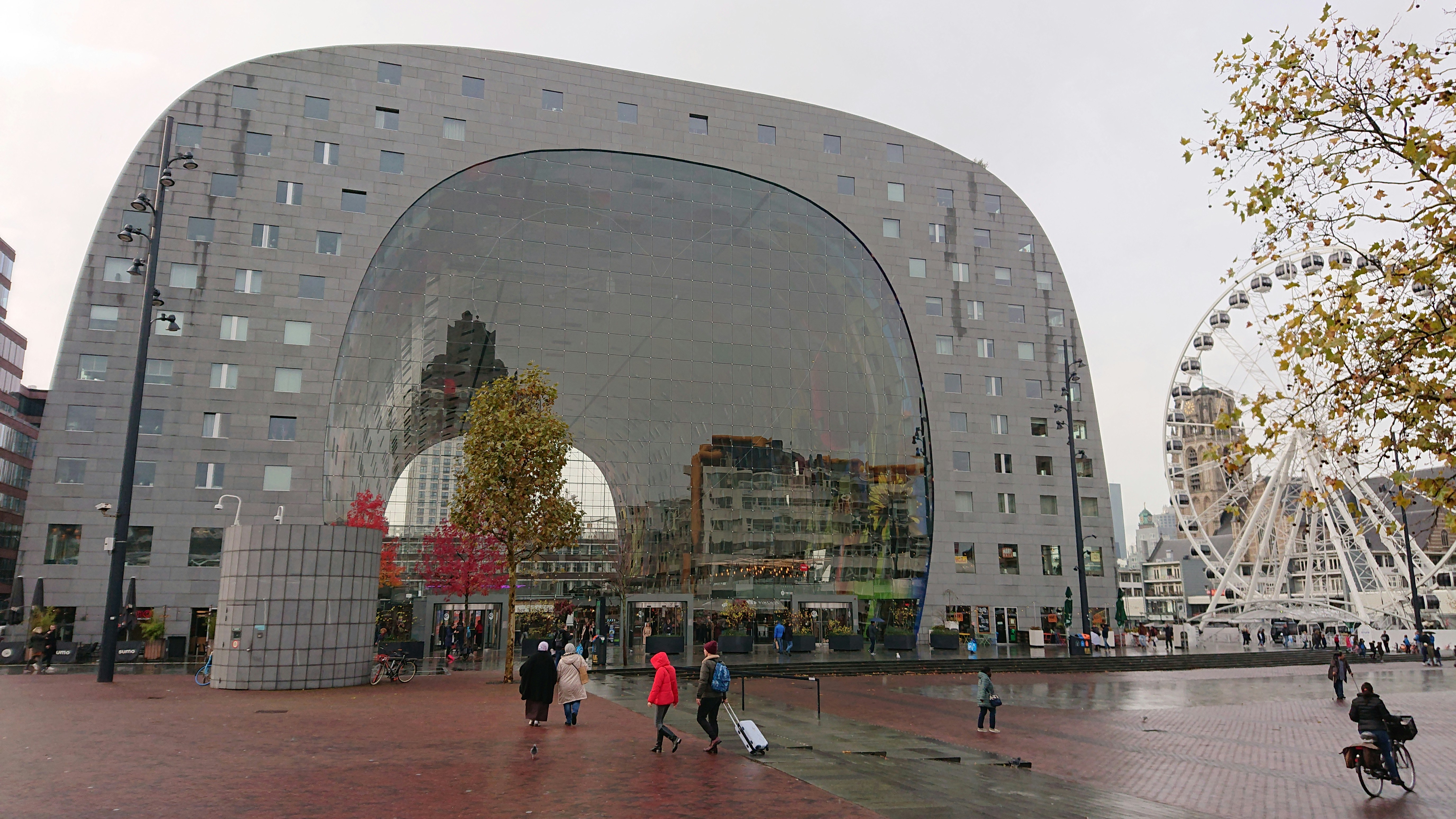 A reflective, contemporary building stands prominently in an urban setting, surrounded by pedestrians and a Ferris wheel in the background.