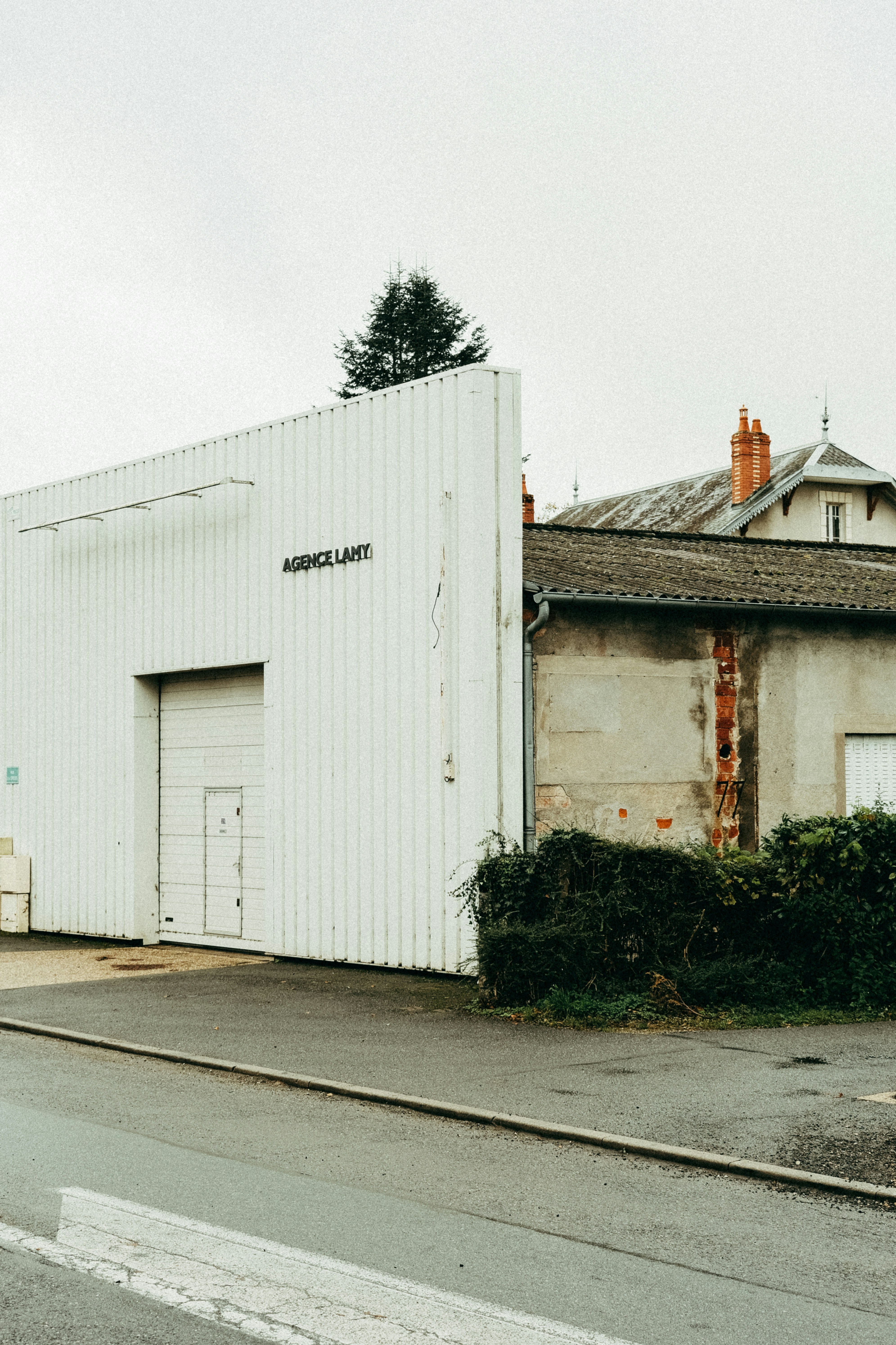 A modern, minimalist building with a white exterior, featuring the name 'AGENCE LANTI' prominently displayed. The structure contrasts with the rustic architecture of the neighboring building.