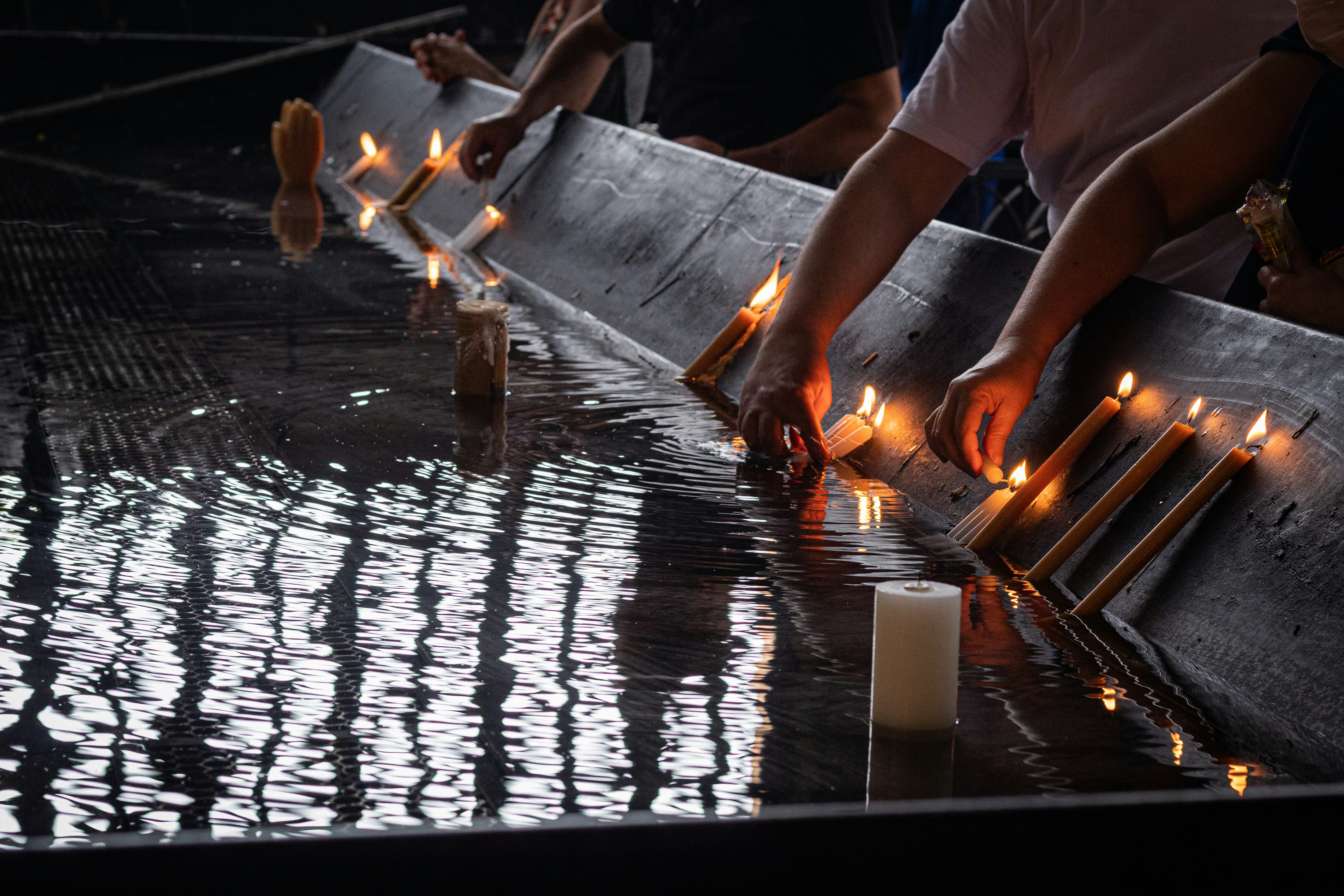 Hands lighting candles along a reflective surface of water, creating a tranquil atmosphere in a spiritual setting.