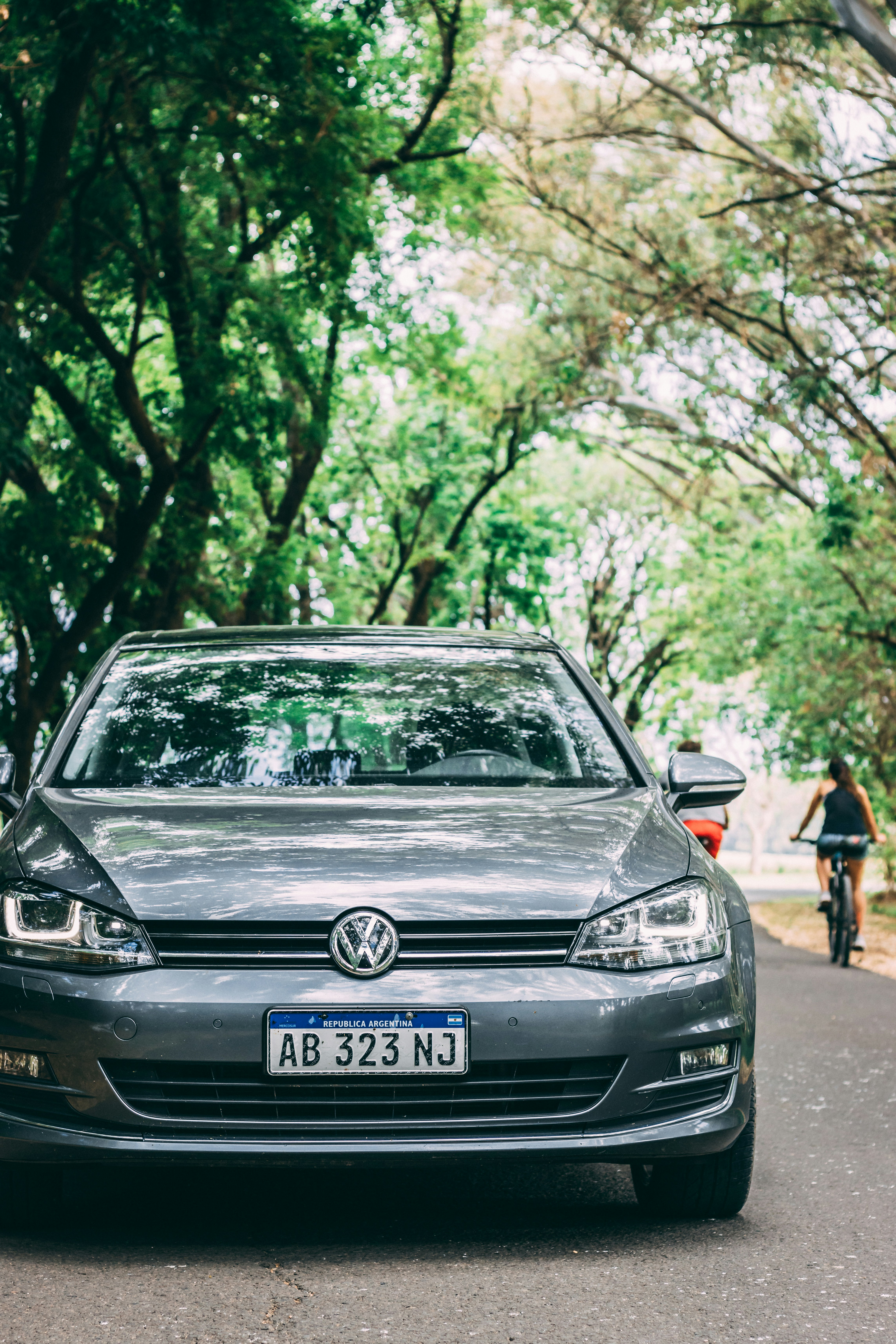 Canopy-Filtered Drive: A VW Front Under Leafy Arches as a Cyclist Passes