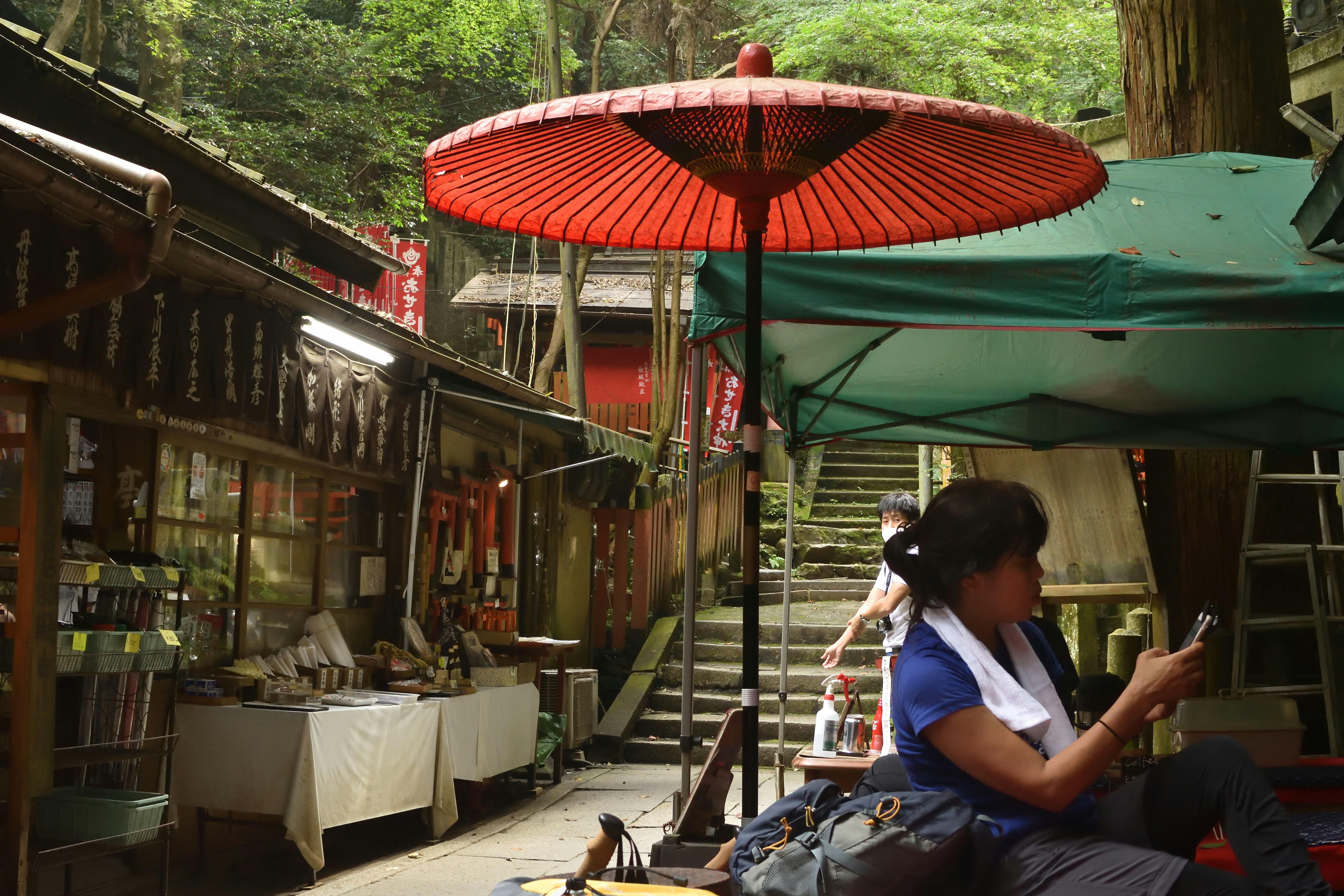 Candid street market scene with stalls along a walkway, a red parasol overhead, and a woman in blue using her phone.