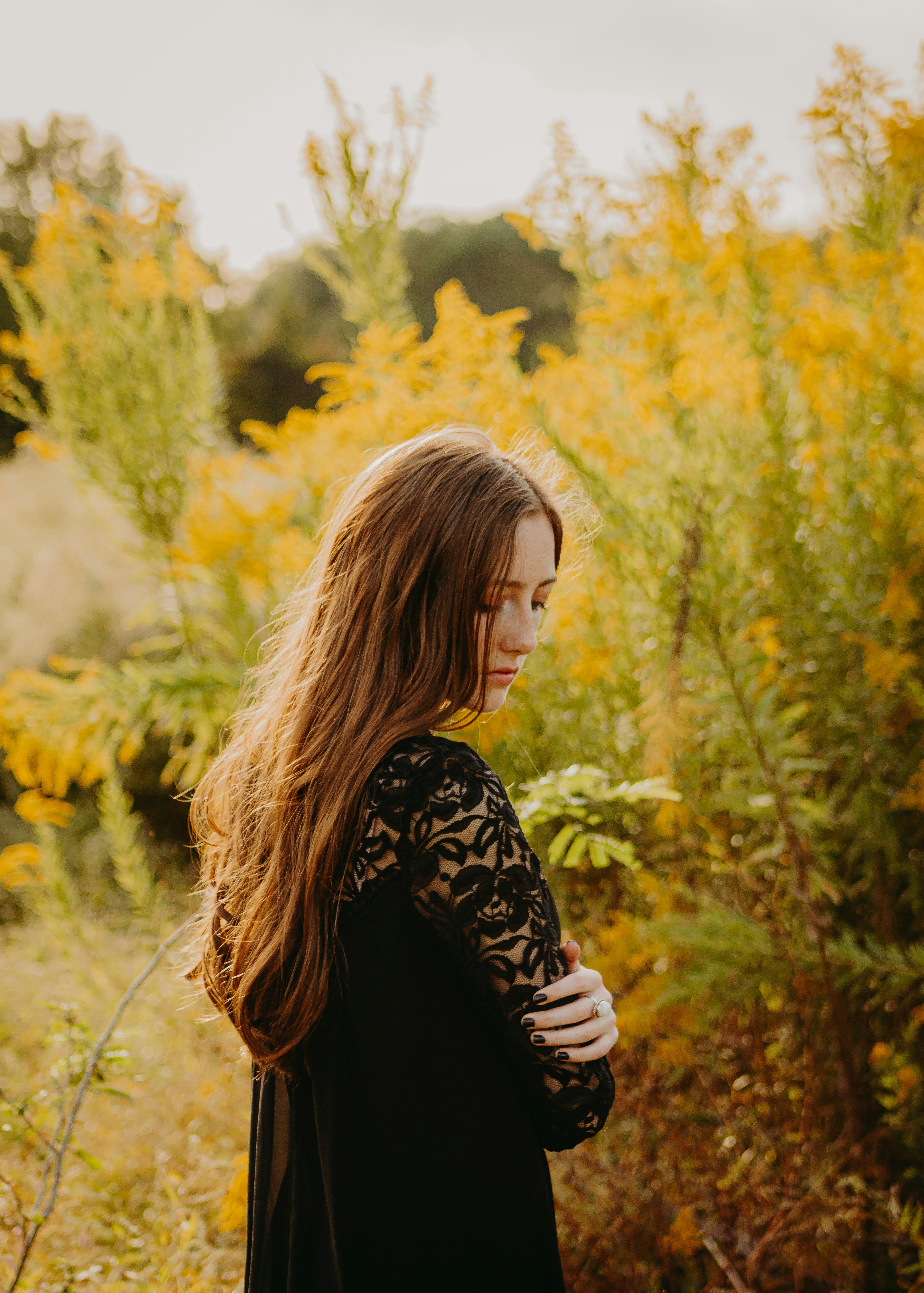 A woman with long, flowing hair stands amidst vibrant yellow foliage, lost in thought. Her black attire contrasts beautifully with the warm autumn colors.