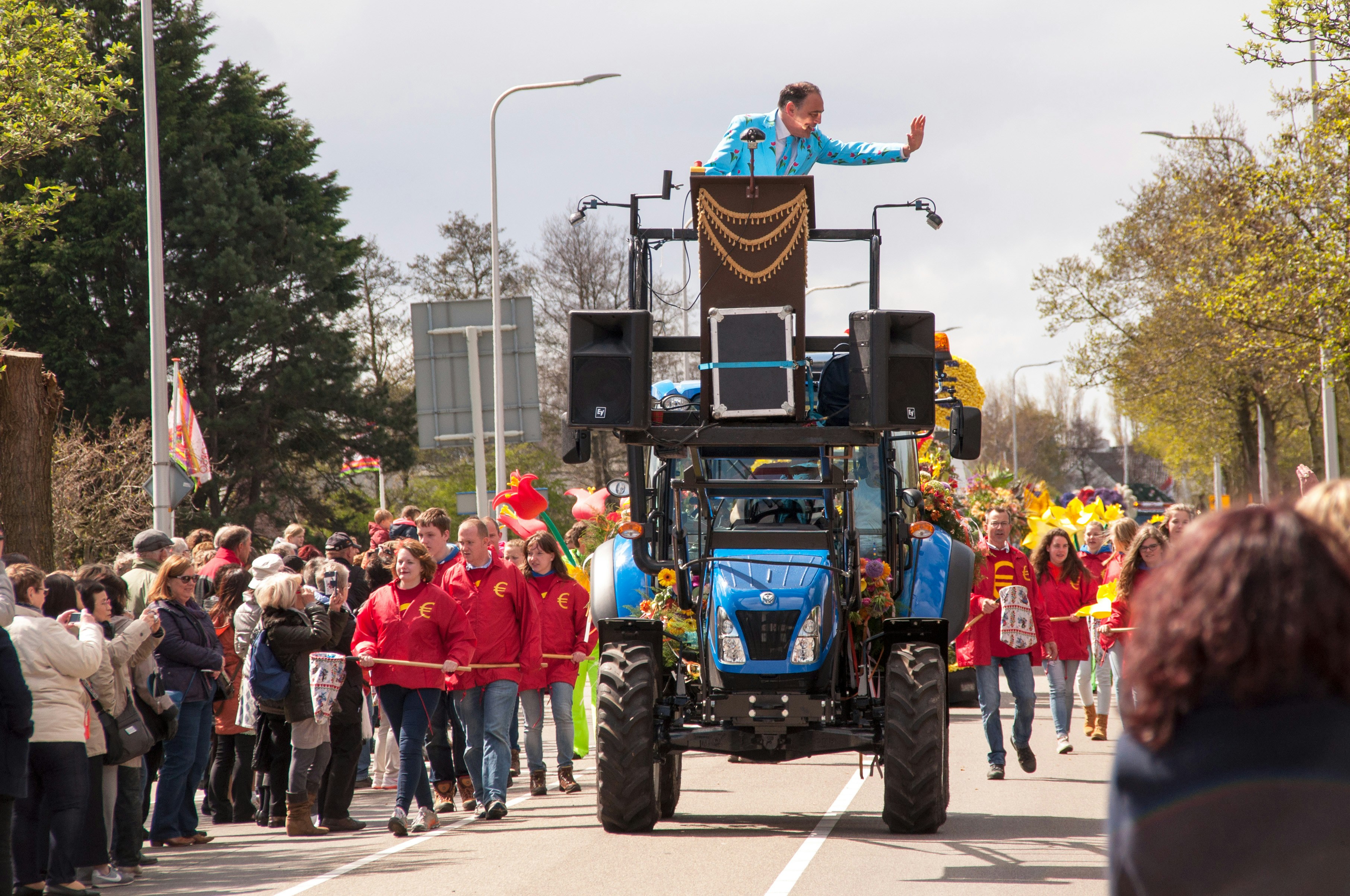 A festive parade featuring a decorated tractor, with a performer waving to the crowd amidst onlookers in red shirts. The scene captures the lively spirit of a community event.