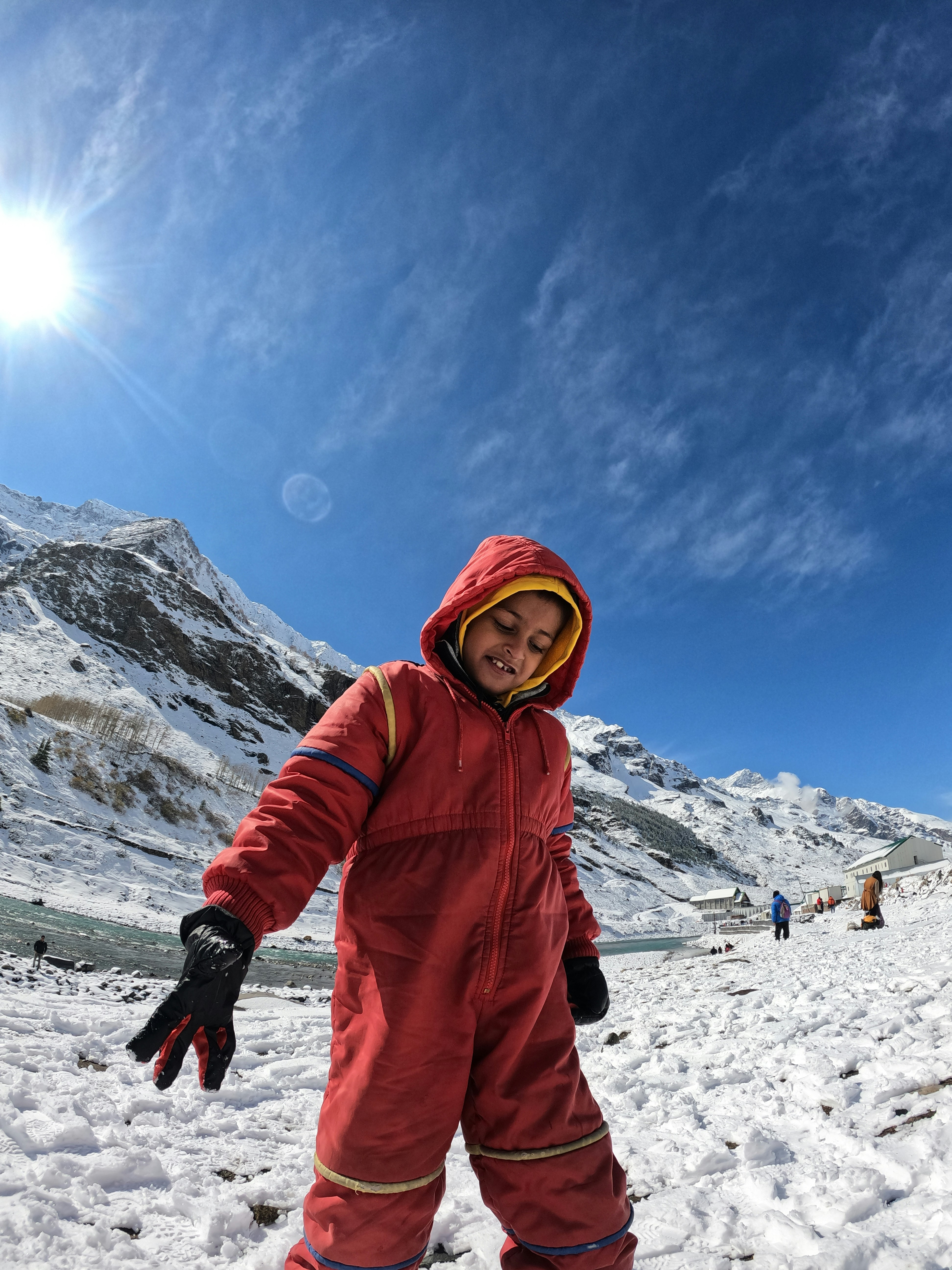 A child in a bright red snowsuit stands on a sunlit snowy slope with rugged alpine peaks under a clear blue sky.