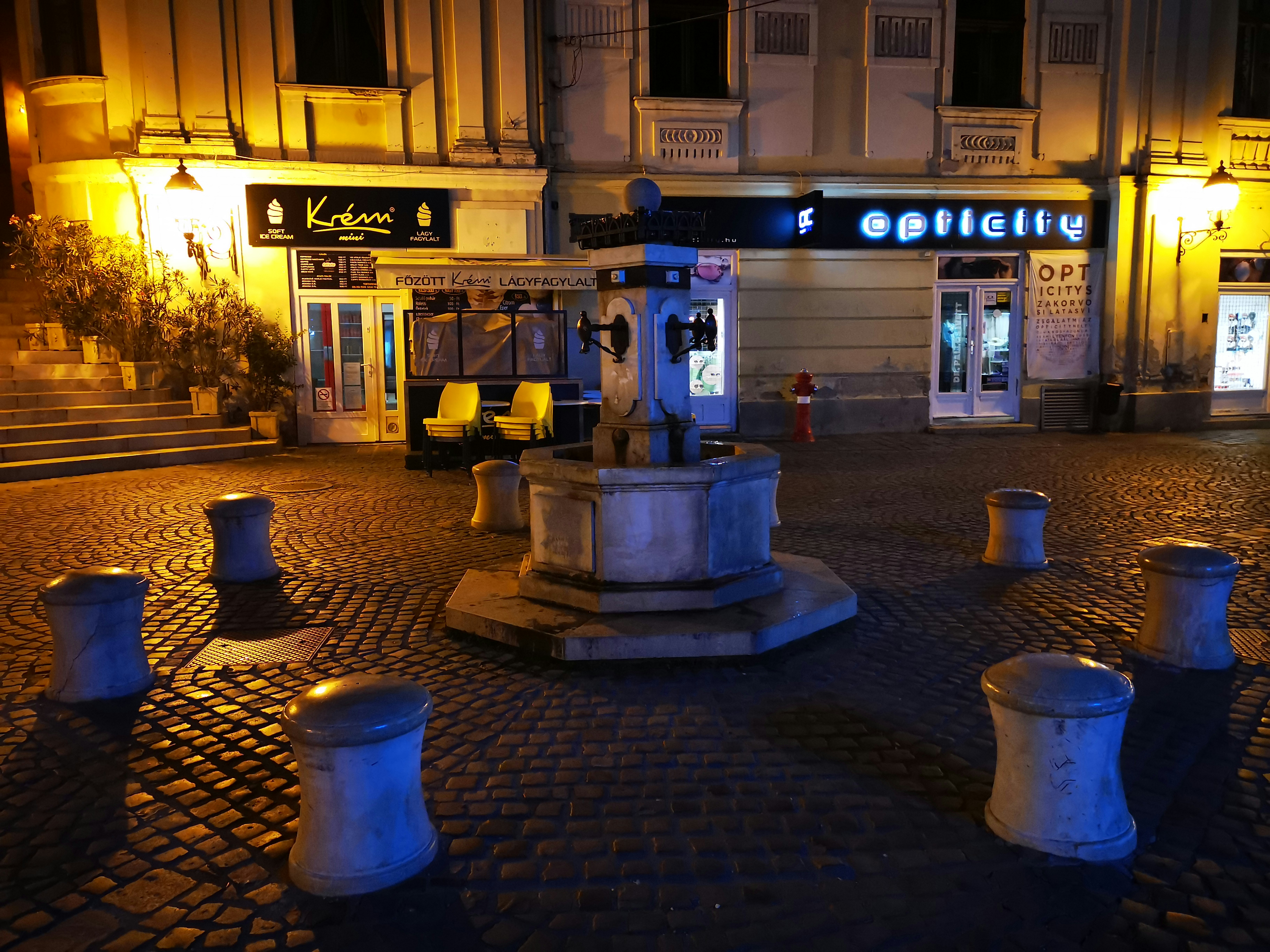 An illuminated fountain stands at the center of a cobblestone square, surrounded by empty seating and softly glowing storefronts, evoking a serene evening atmosphere.