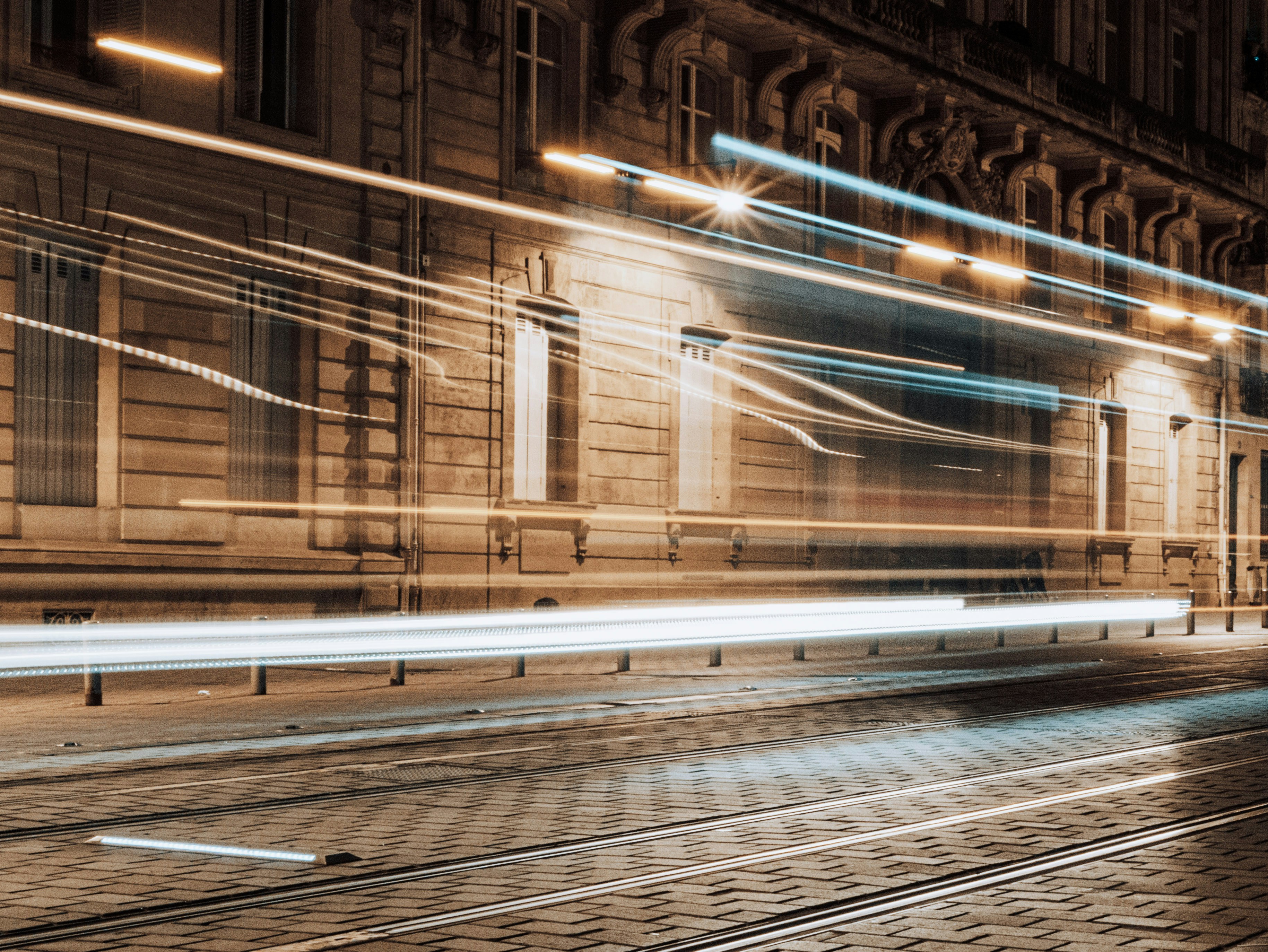 Streaks of light from passing trams illuminate the cobblestone street, showcasing the energy of a bustling city at night.