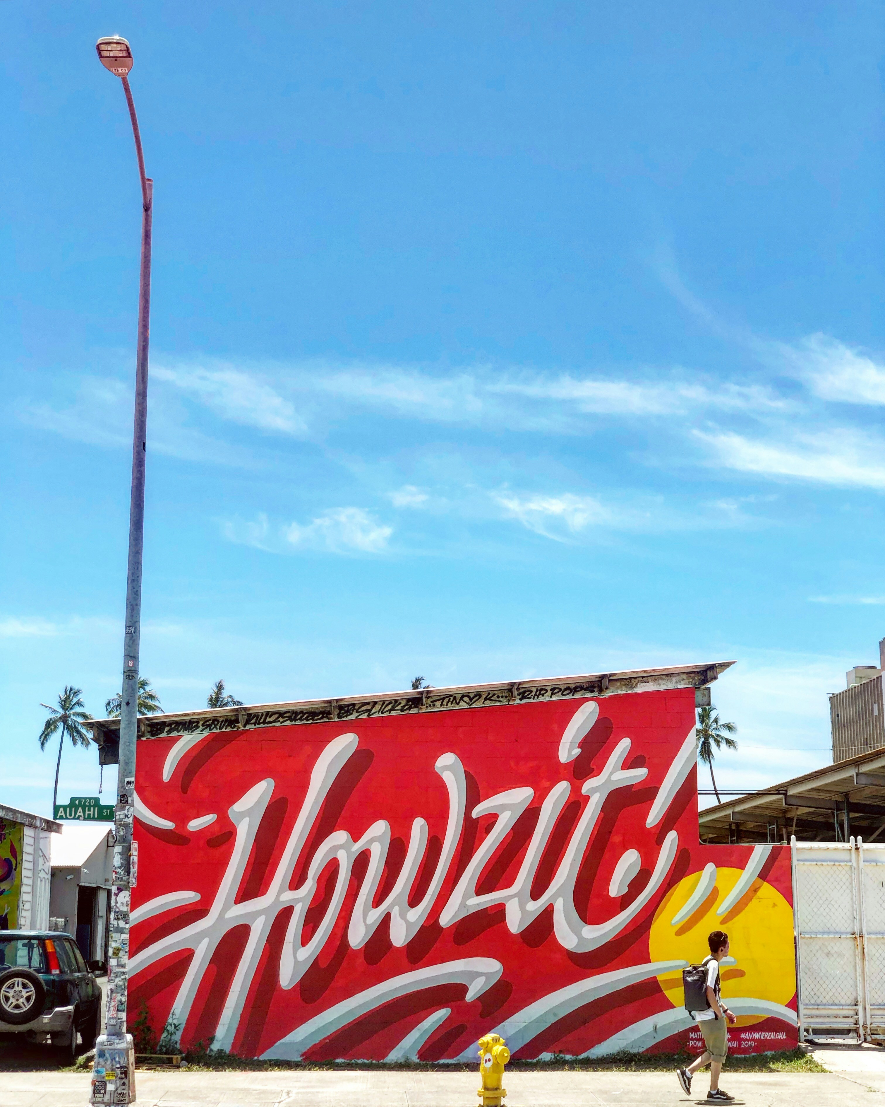 Bold mural featuring the word 'Howzit' against a bright red background, complemented by a sunny blue sky. A pedestrian and a parked car add life to the urban scene.