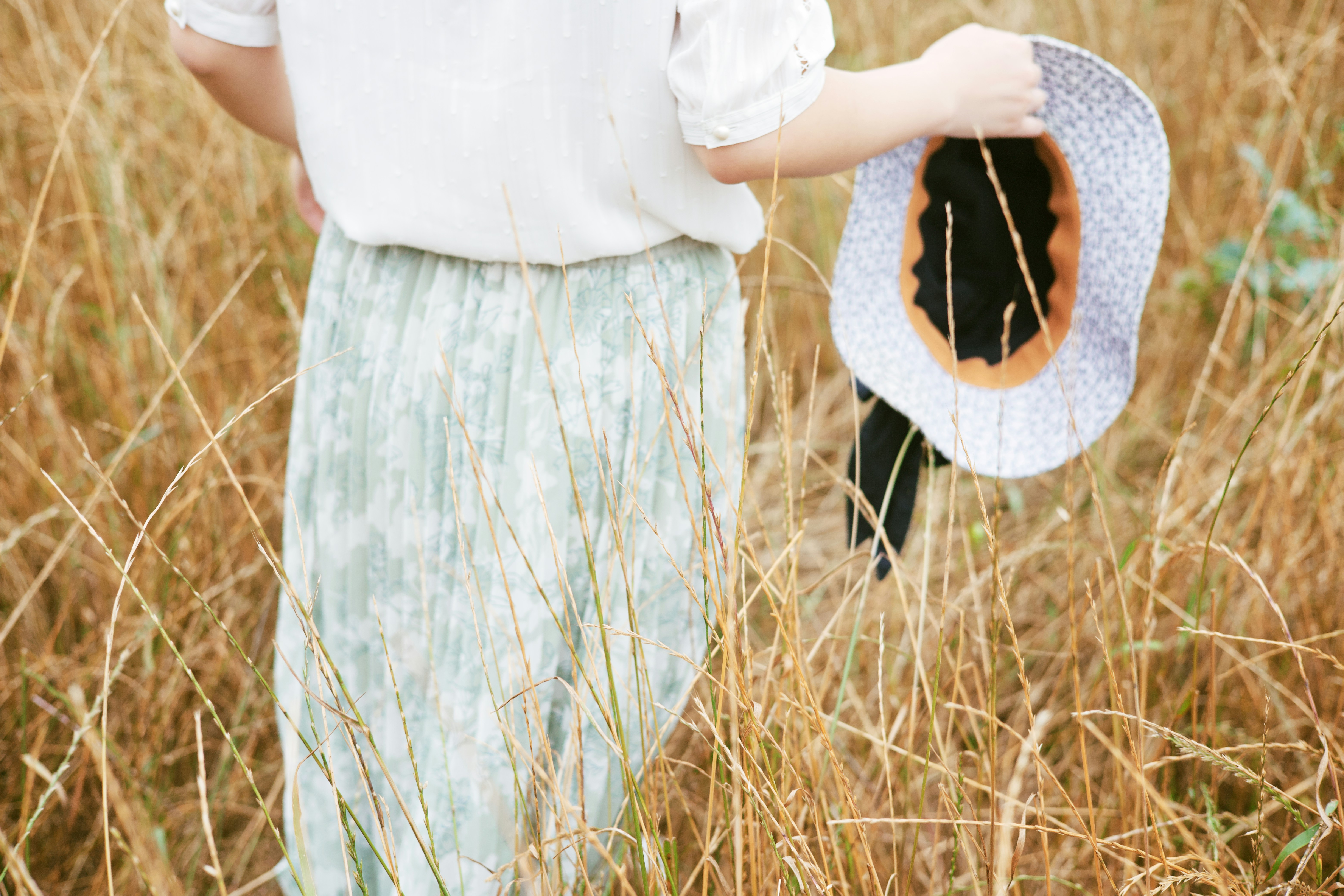 A person in a light blouse and patterned skirt holds a sun hat while walking through tall golden grass. The scene evokes a sense of nostalgia and tranquility.