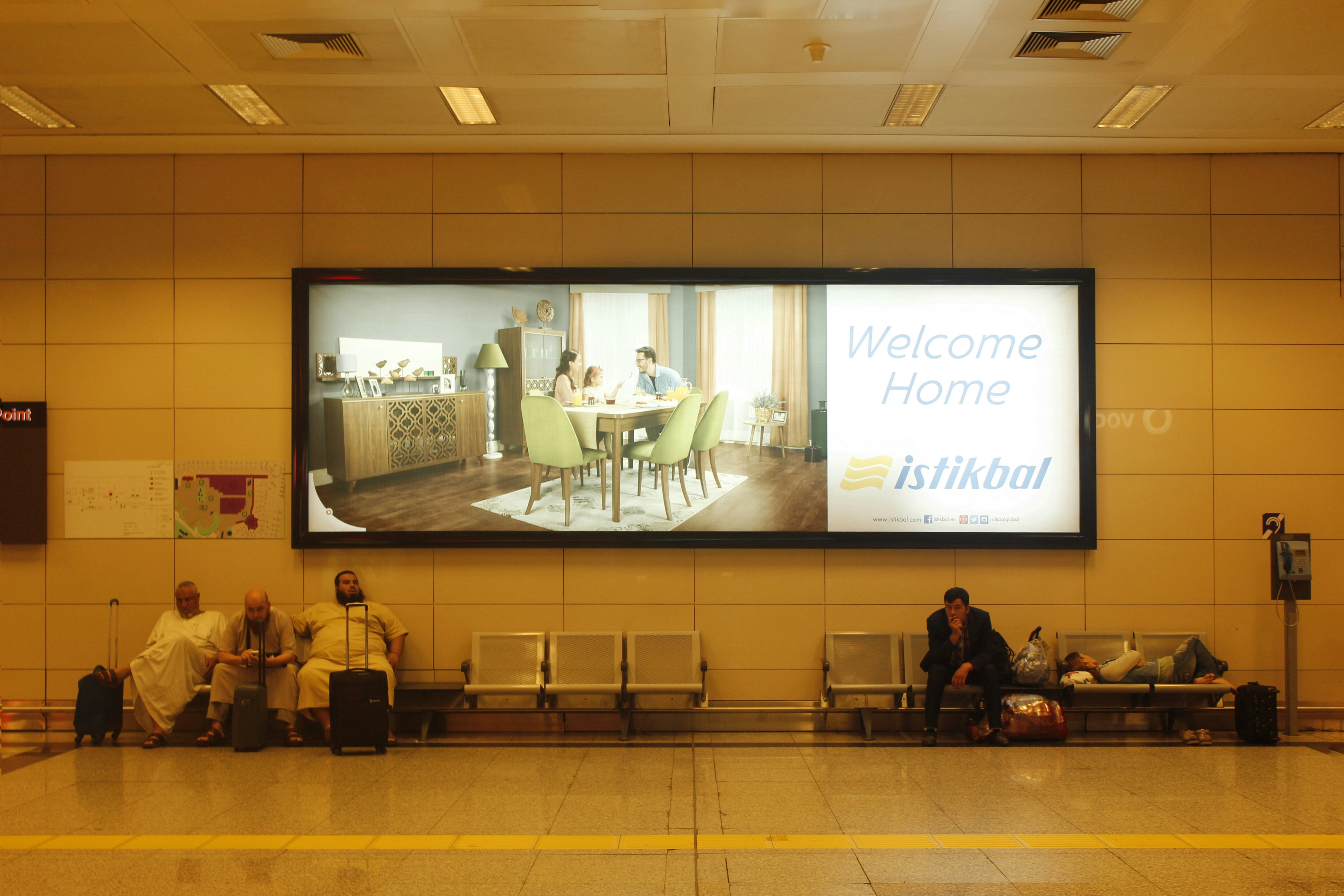 Passengers resting in an airport terminal with a large advertisement displaying a welcoming message and homey scene.