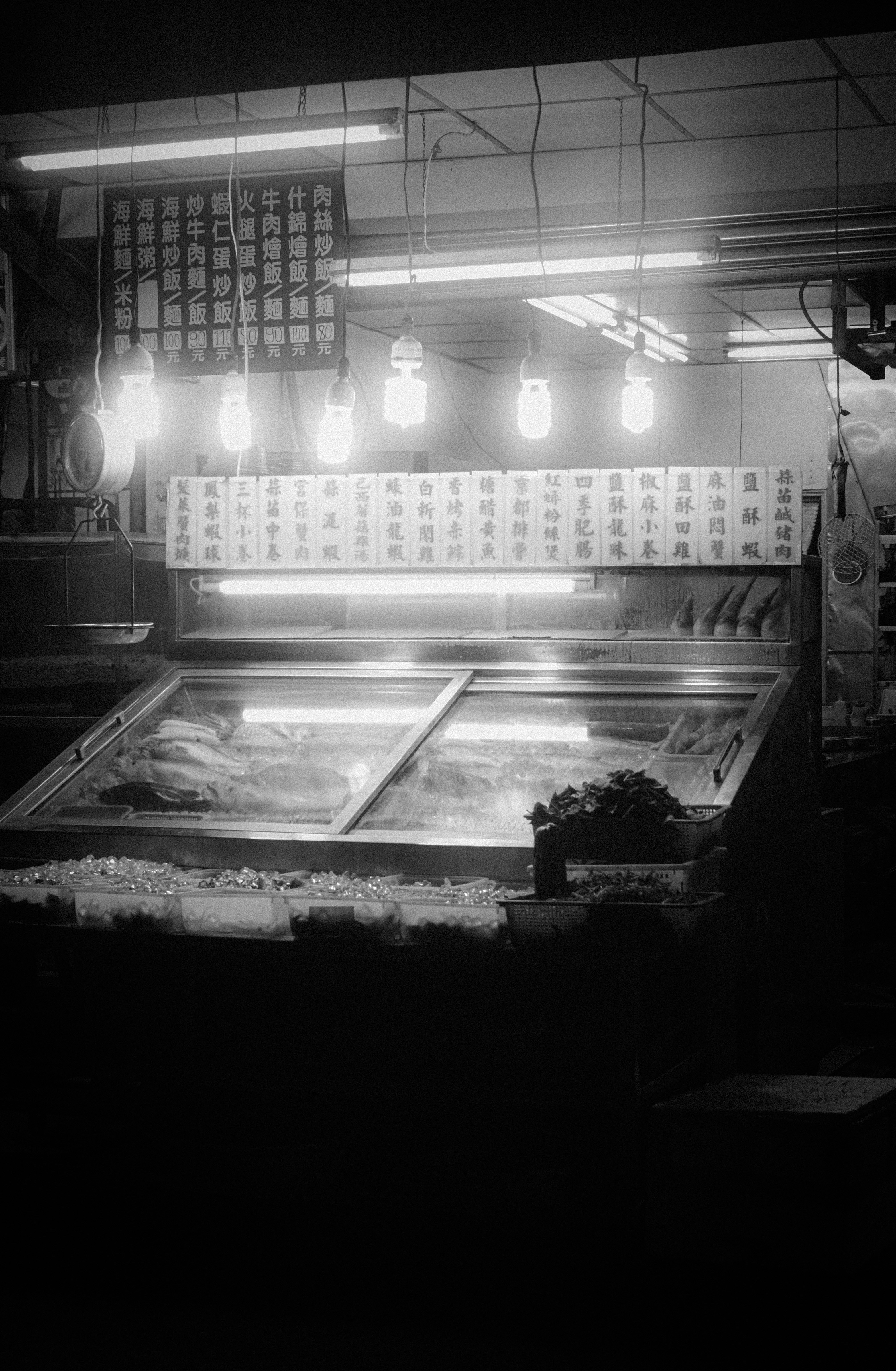 Dimly lit fish market stall showcasing fresh seafood under glowing lights, with handwritten menus above. A hint of greenery adds a touch of freshness.