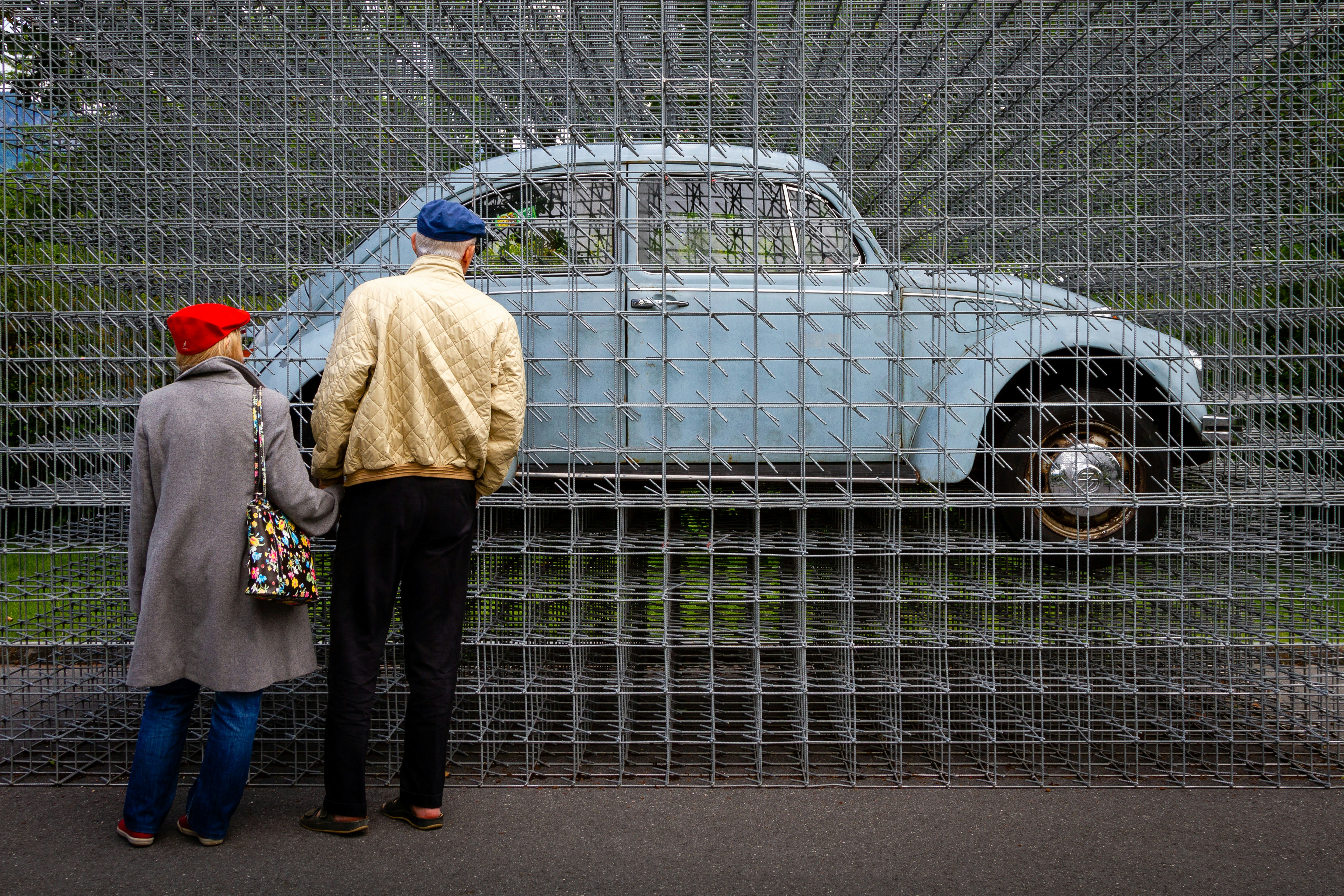 Two Elder Observers Gaze at a Dim Vintage Car Behind Chainlink